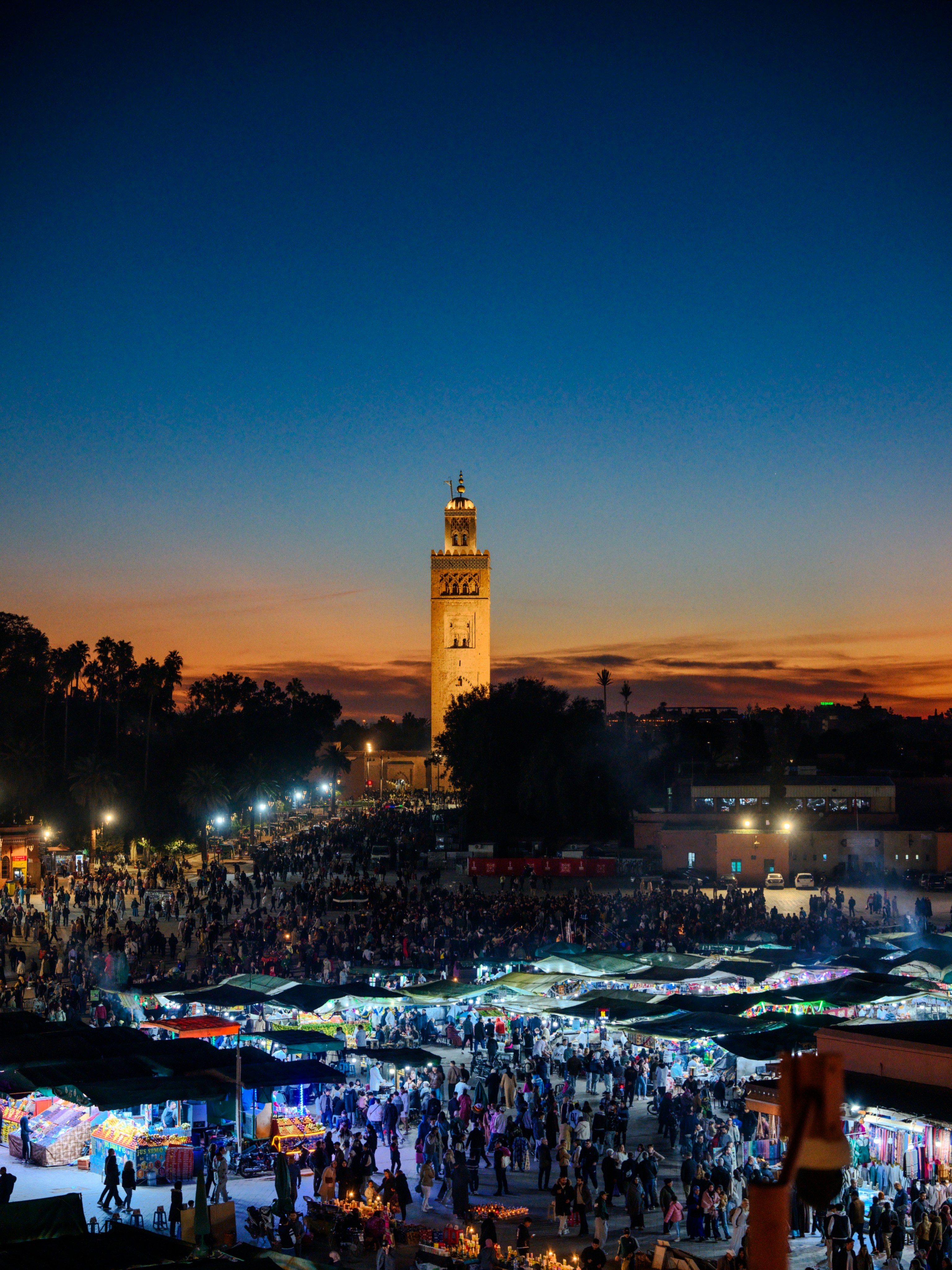 Night falls over Marrakesh and the city comes fully alive. Jemaa el-Fnaa spreads out below in a sea of glowing market stalls, storytellers, food vendors, musicians, and the constant flow of people moving through light and shadow. In the distance, the illuminated minaret of the Koutoubia Mosque rises calmly above the palm trees, anchoring the chaos with centuries of history. The sky fades from deep blue to warm orange, framing the square as smoke, sound, and color blend into one unmistakable rhythm. This is Marrakesh after dark. Loud, layered, timeless, and impossible to ignore.