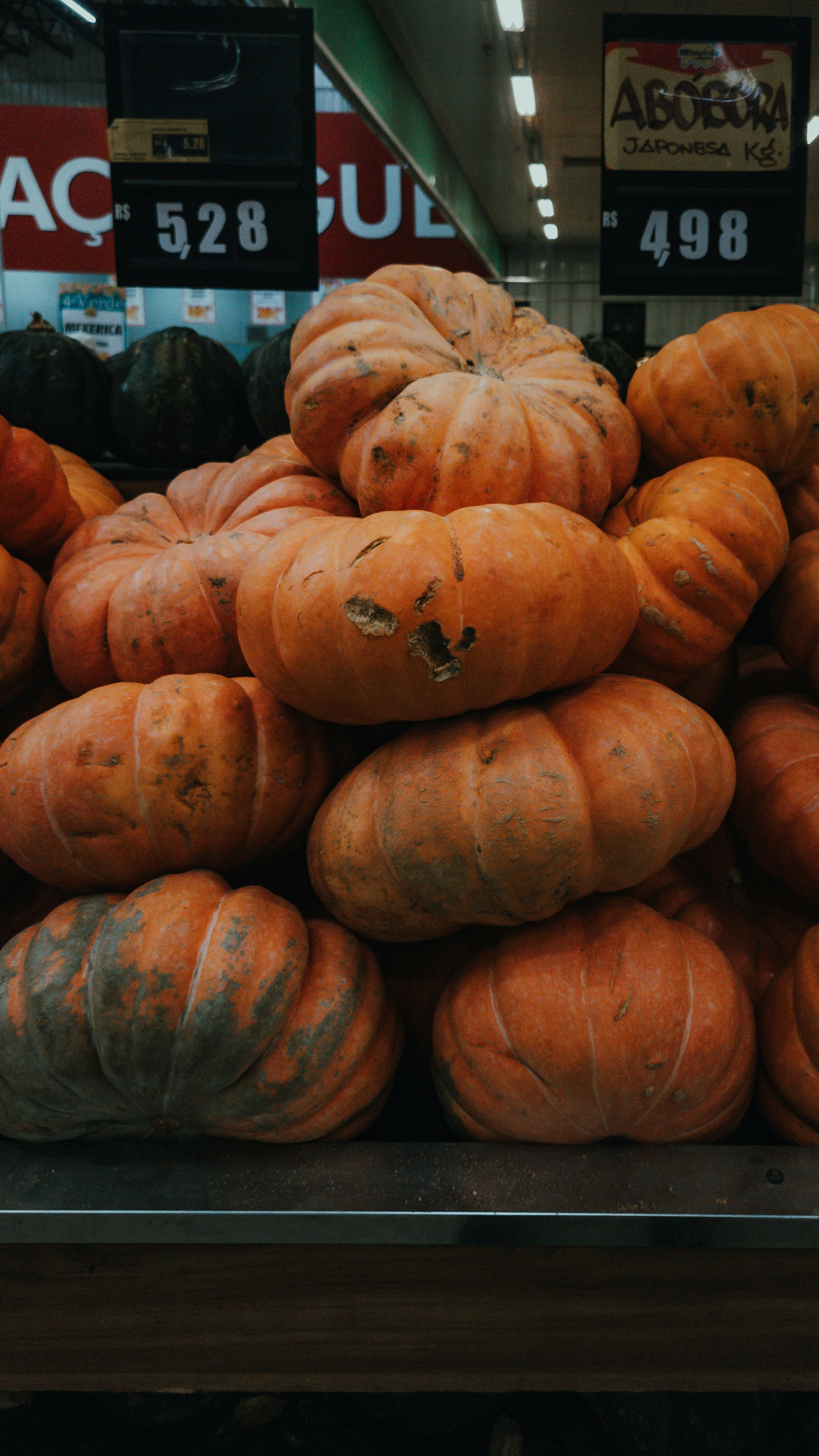 A dense mound of orange pumpkins stacked at a market stall with price signs in the background; the scene emphasizes harvest season and retail display.