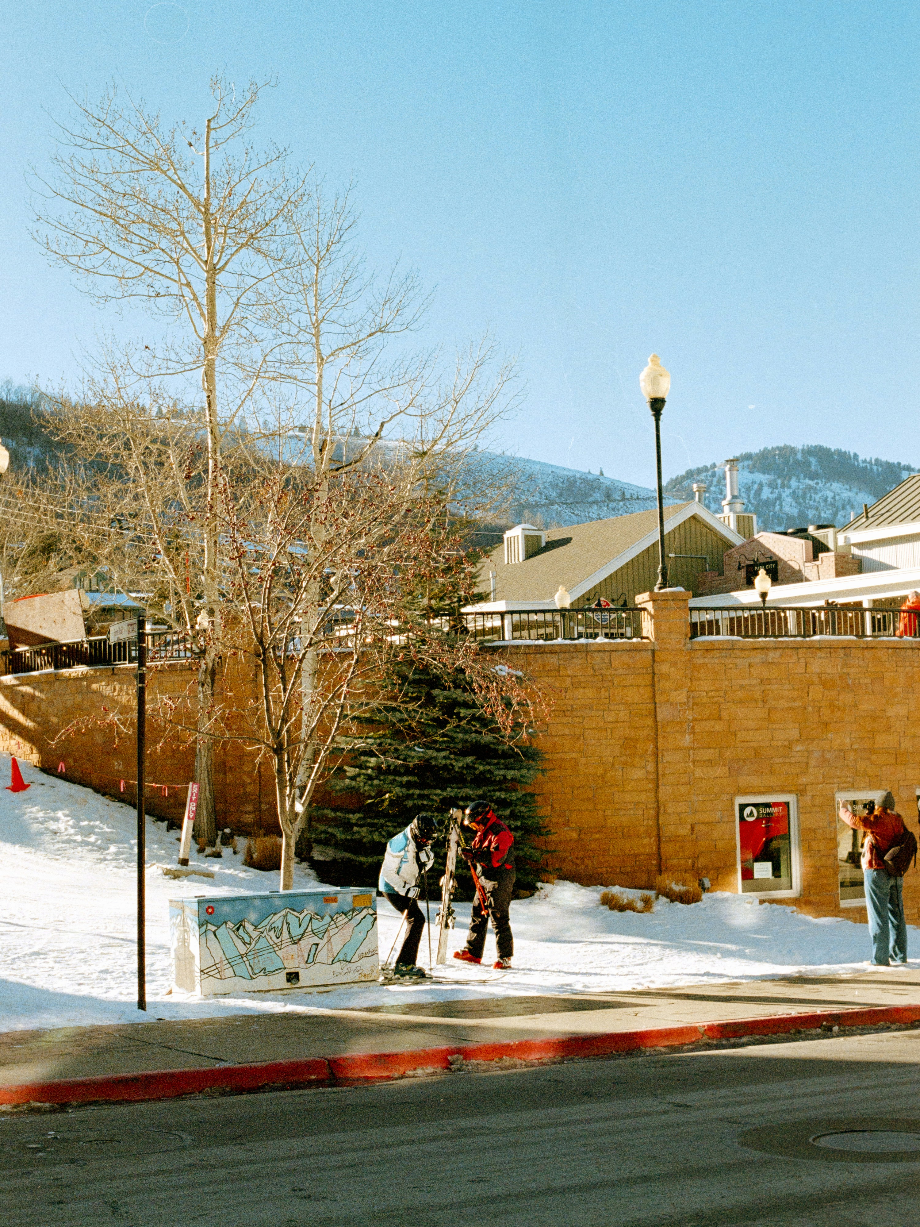 Skiers practice on a snowy slope beside a brick wall and a lamppost with a distant hillside; a small group observes near a utility box and a mural. This is a candid street scene with real-world activity.