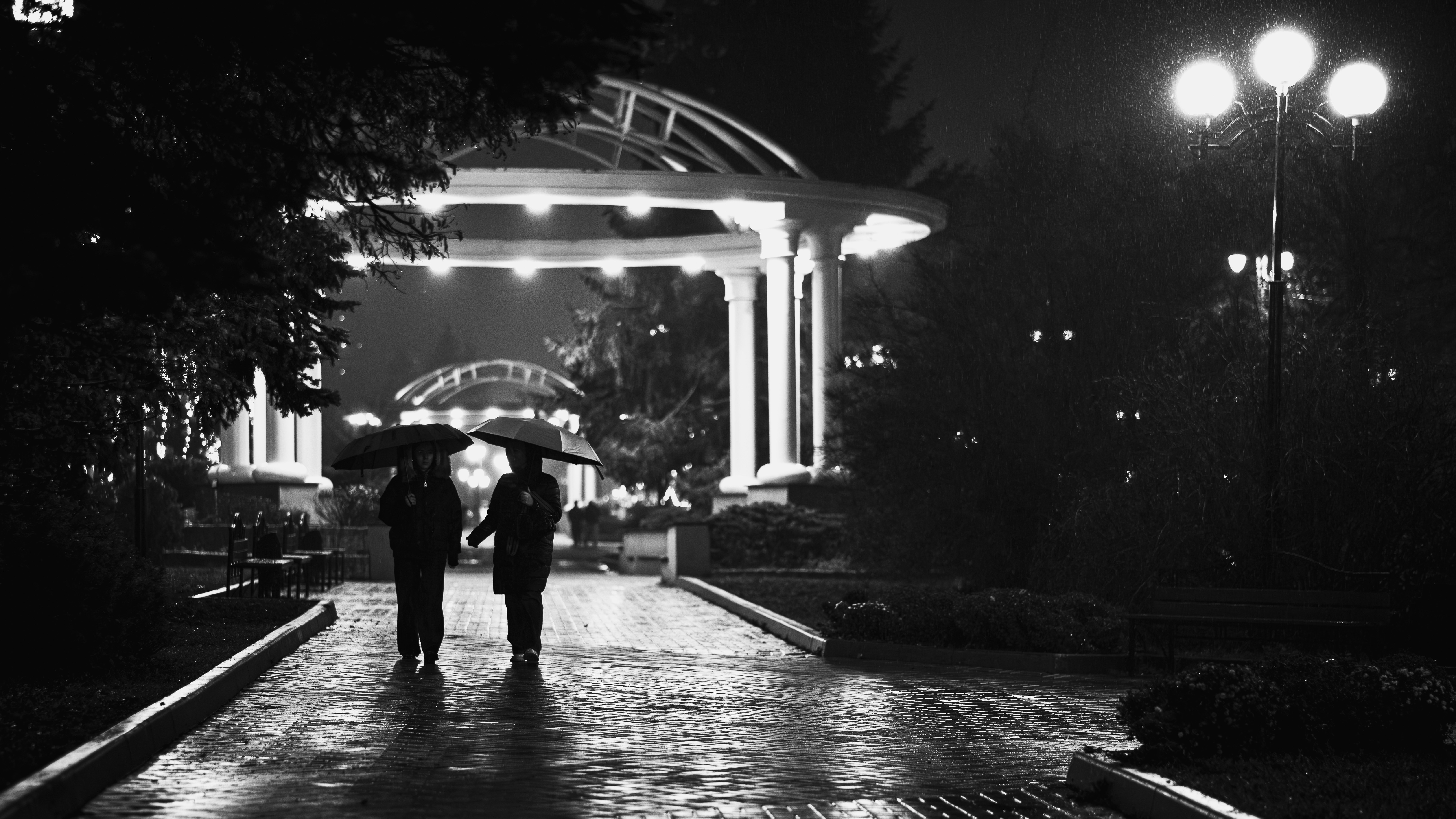 Two Figures under Neon Archway: Nighttime Rain on the Plaza