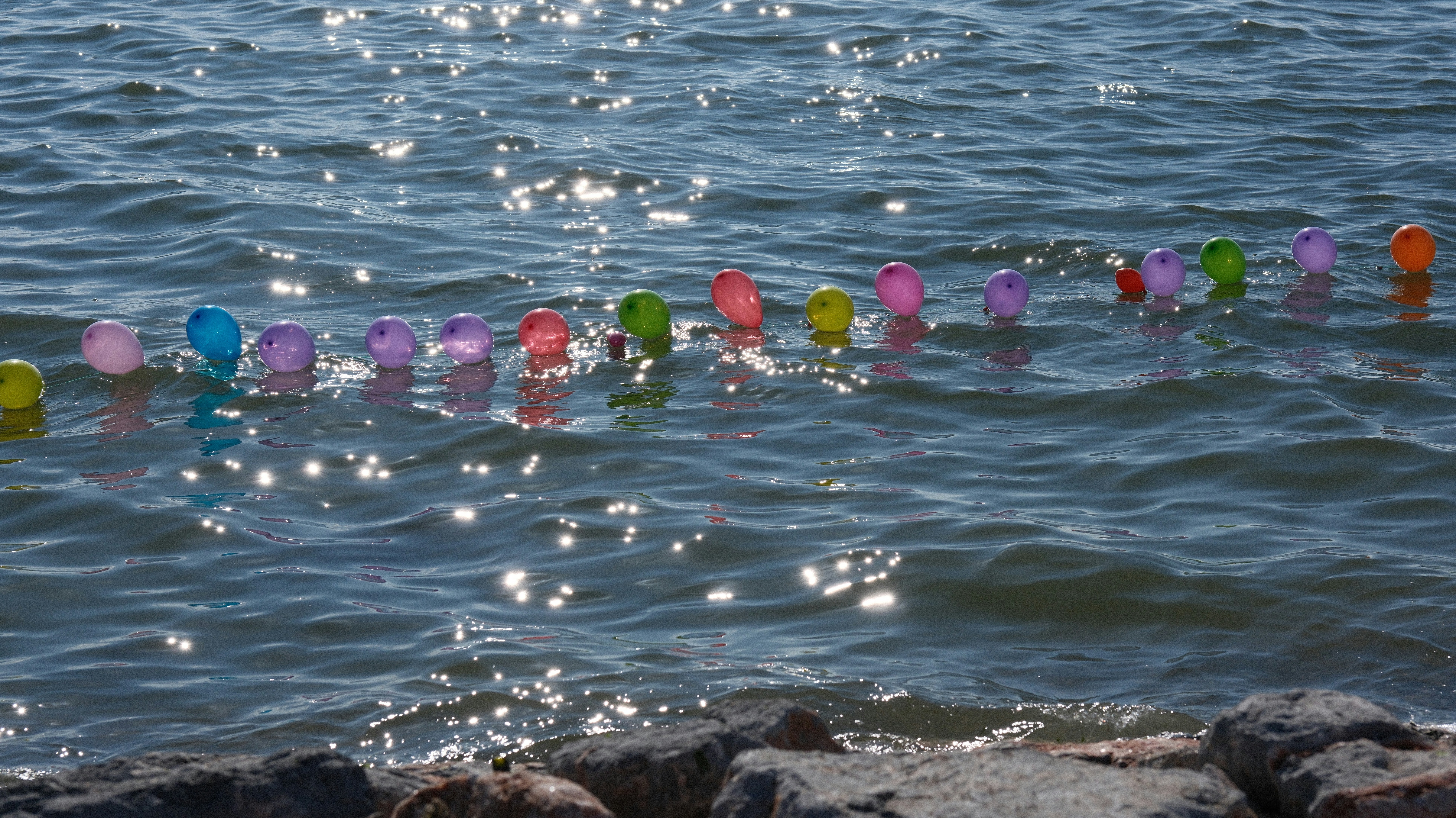 Colorful balloons float along a shimmering sea line, tethered together as gentle waves lap the rocky shore. This appears as a decorative installation or event display over water.