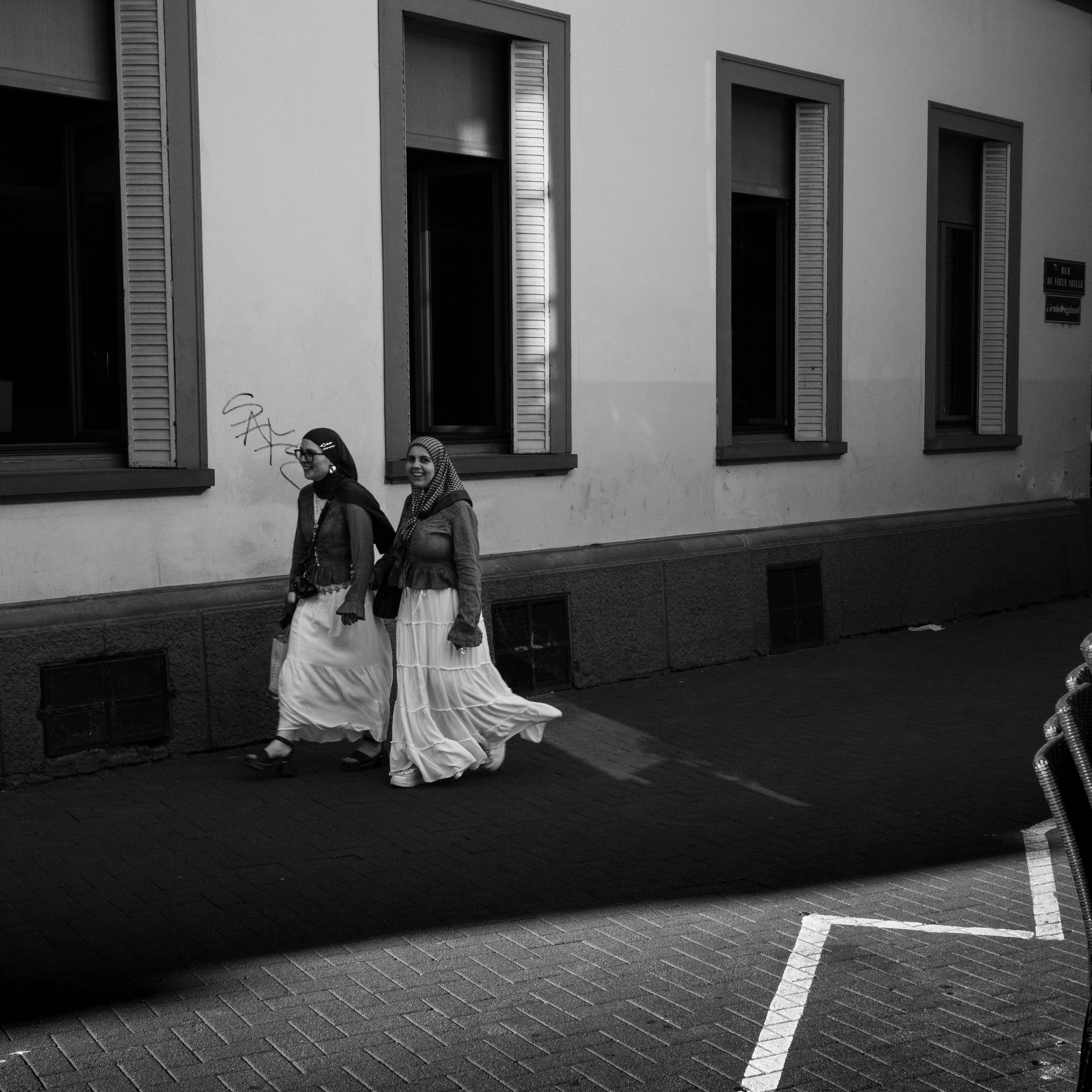 Two Women in Long White Skirts Walk Past a Graffitied Wall