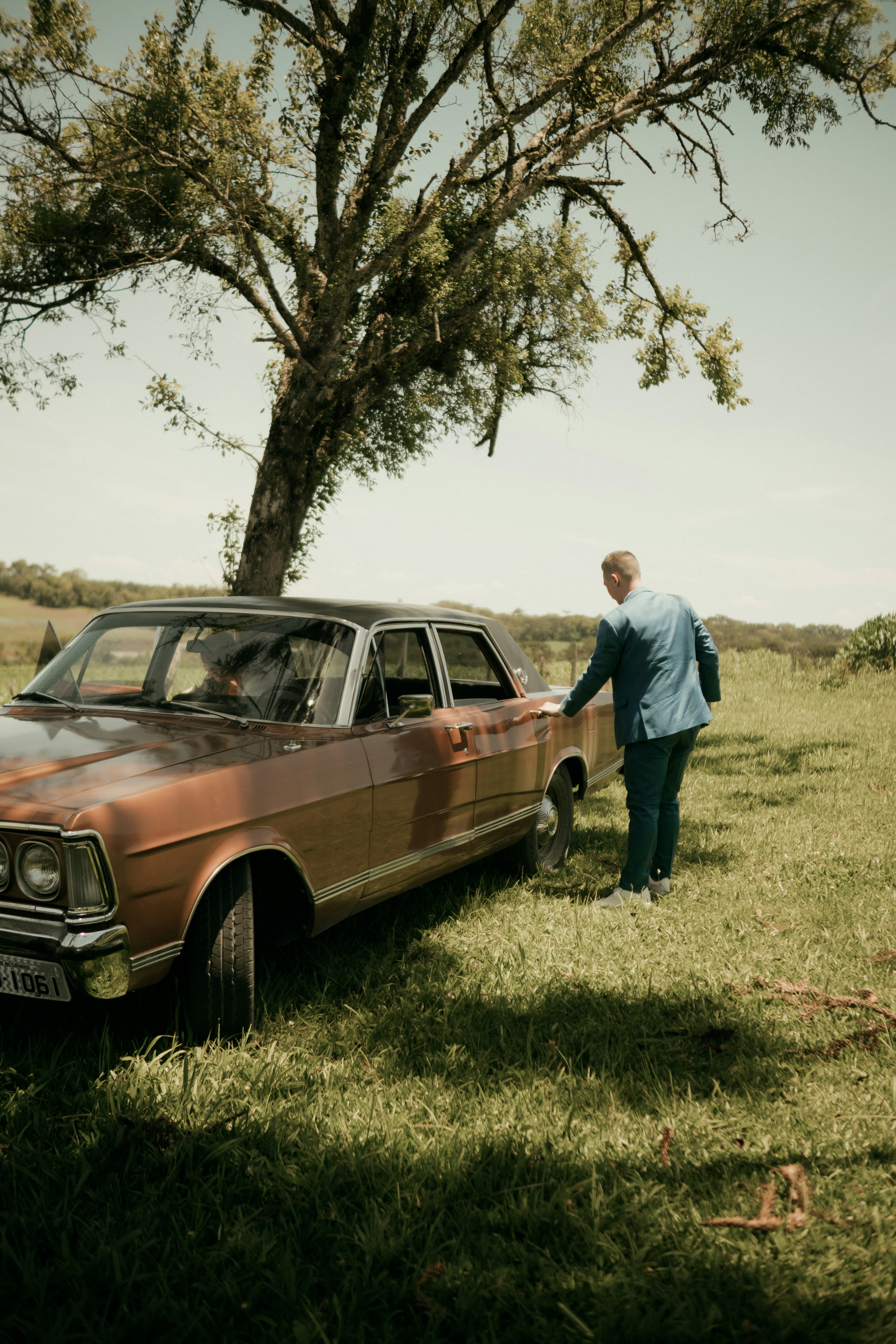 Vintage Encounter: A Man in a Blue Jacket Approaches a Classic Car Under a Spreading Tree