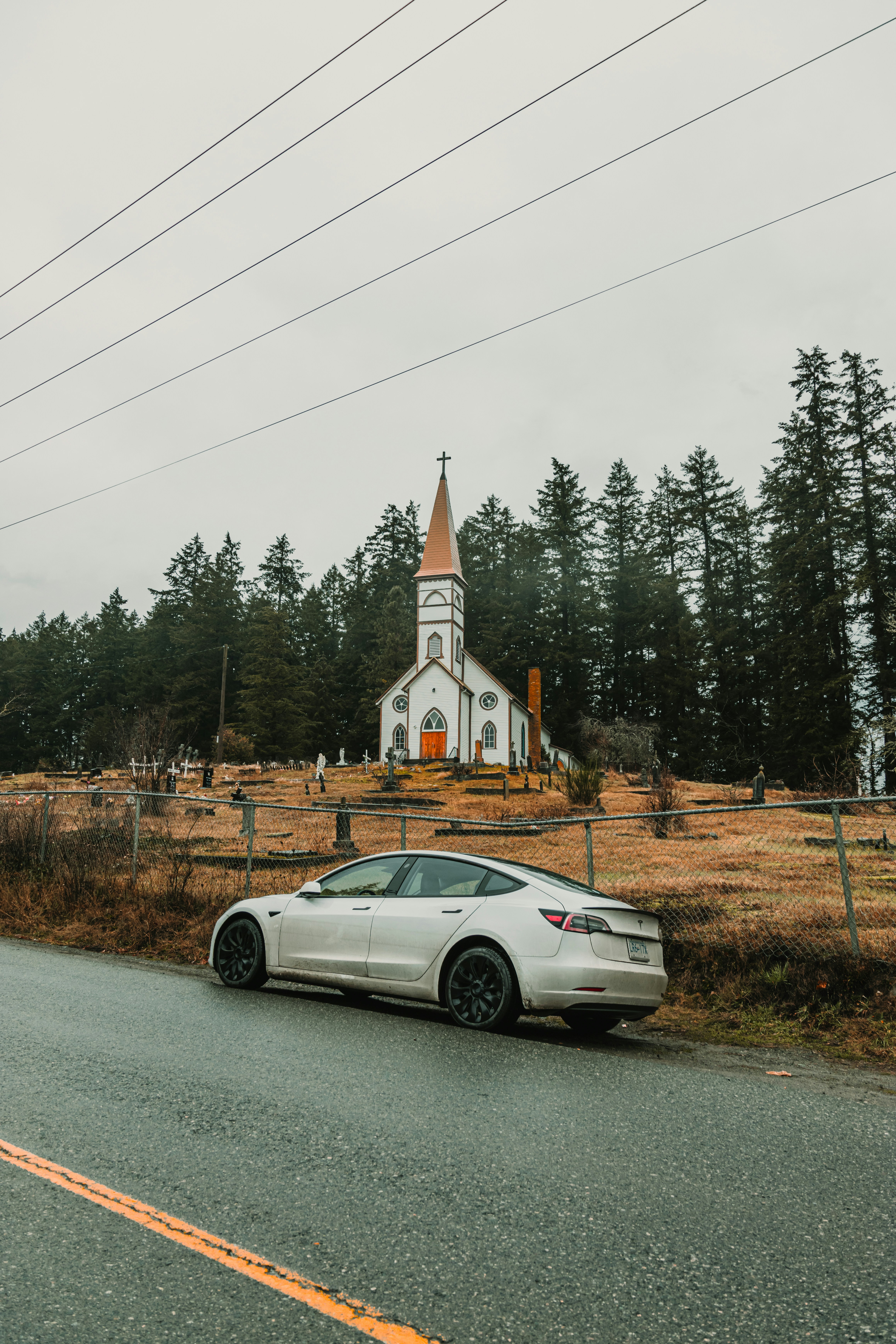 White Tesla by a hillside chapel under overcast skies