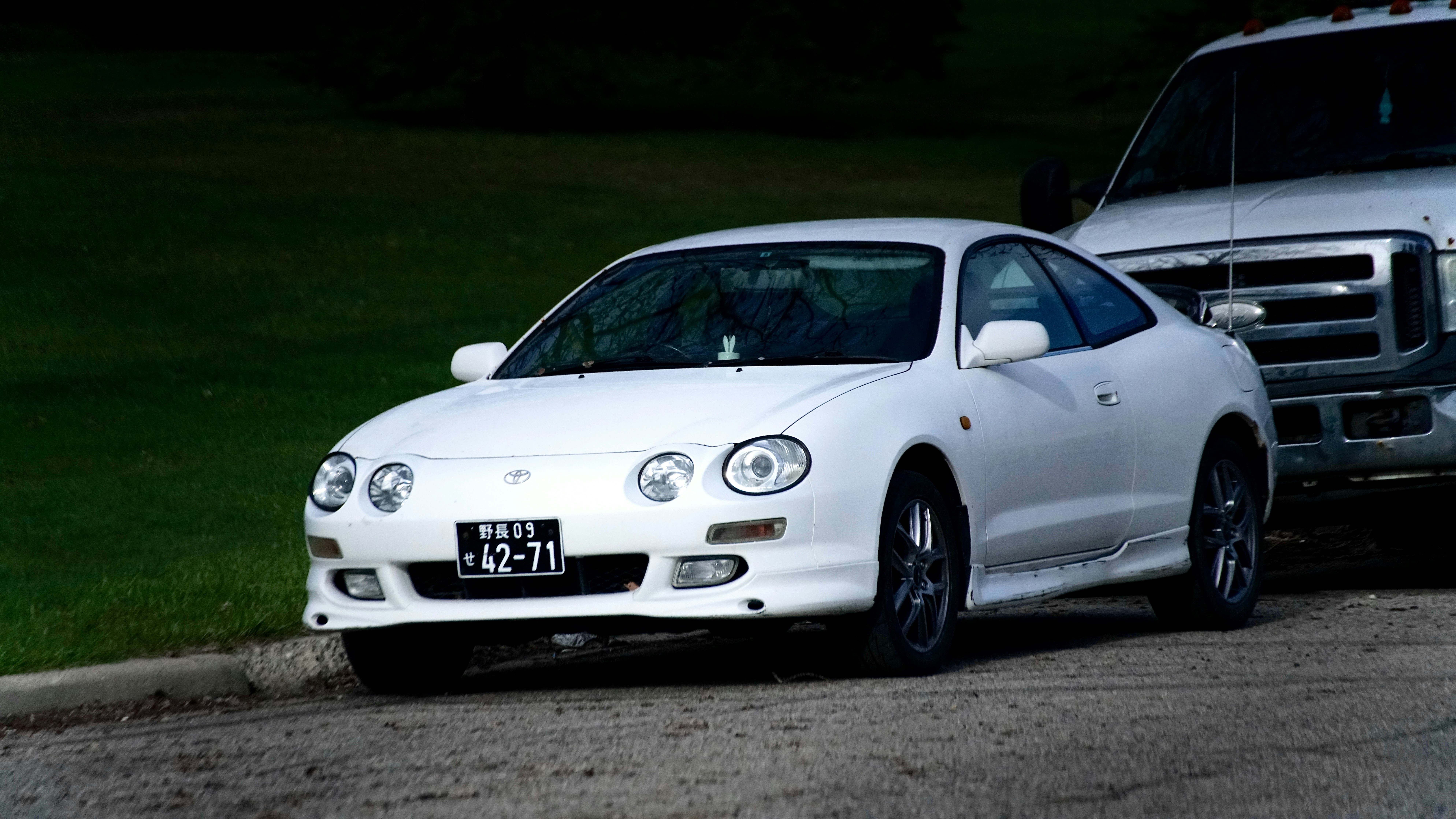 White sports coupe crossing a parkway, shadowed by a heavy-duty truck
