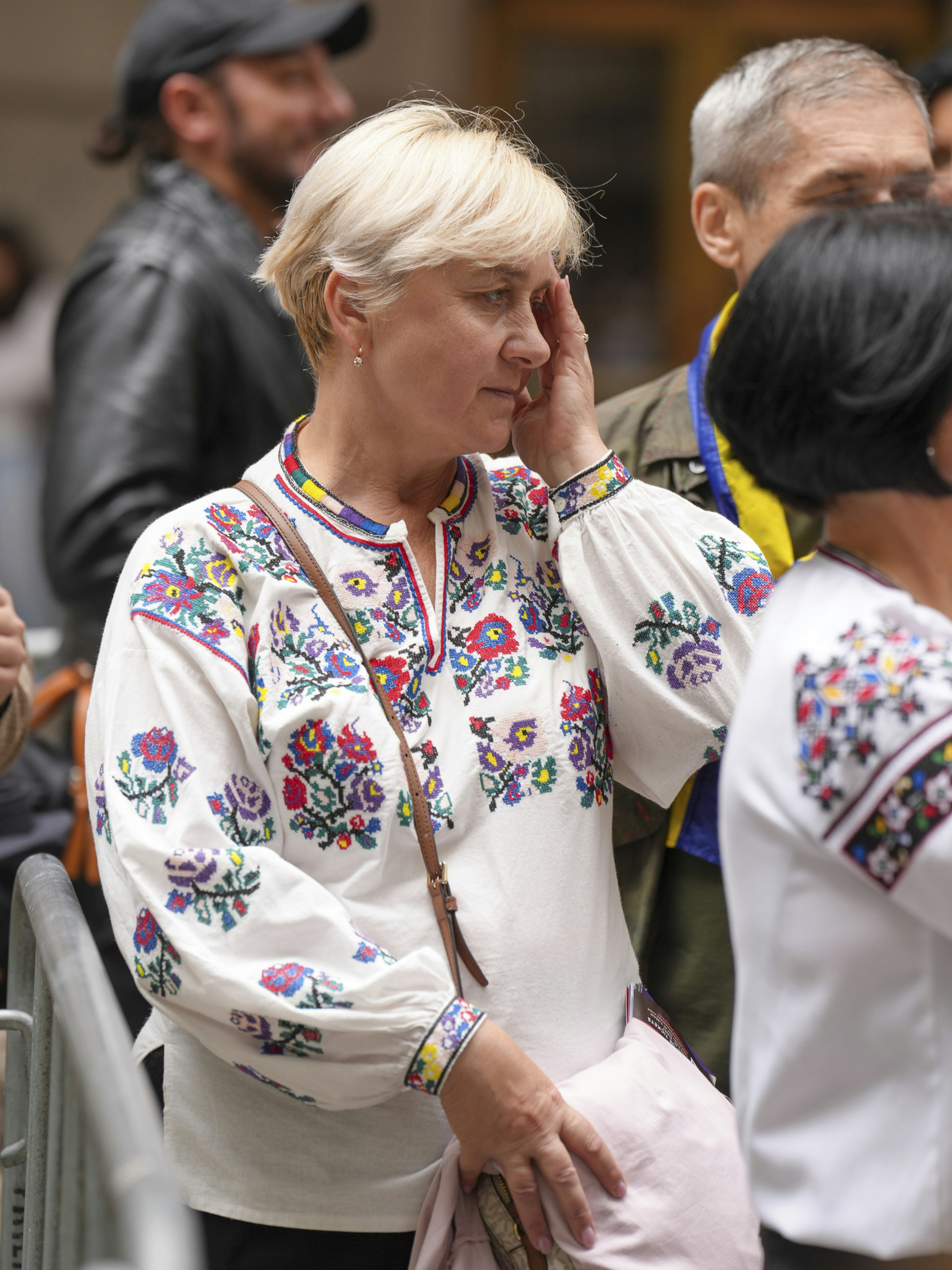 A woman in a white embroidered blouse stands in a crowd, her hand raised to her face in a thoughtful pause, while others bustle behind. The scene captures traditional textile details and candid street life.