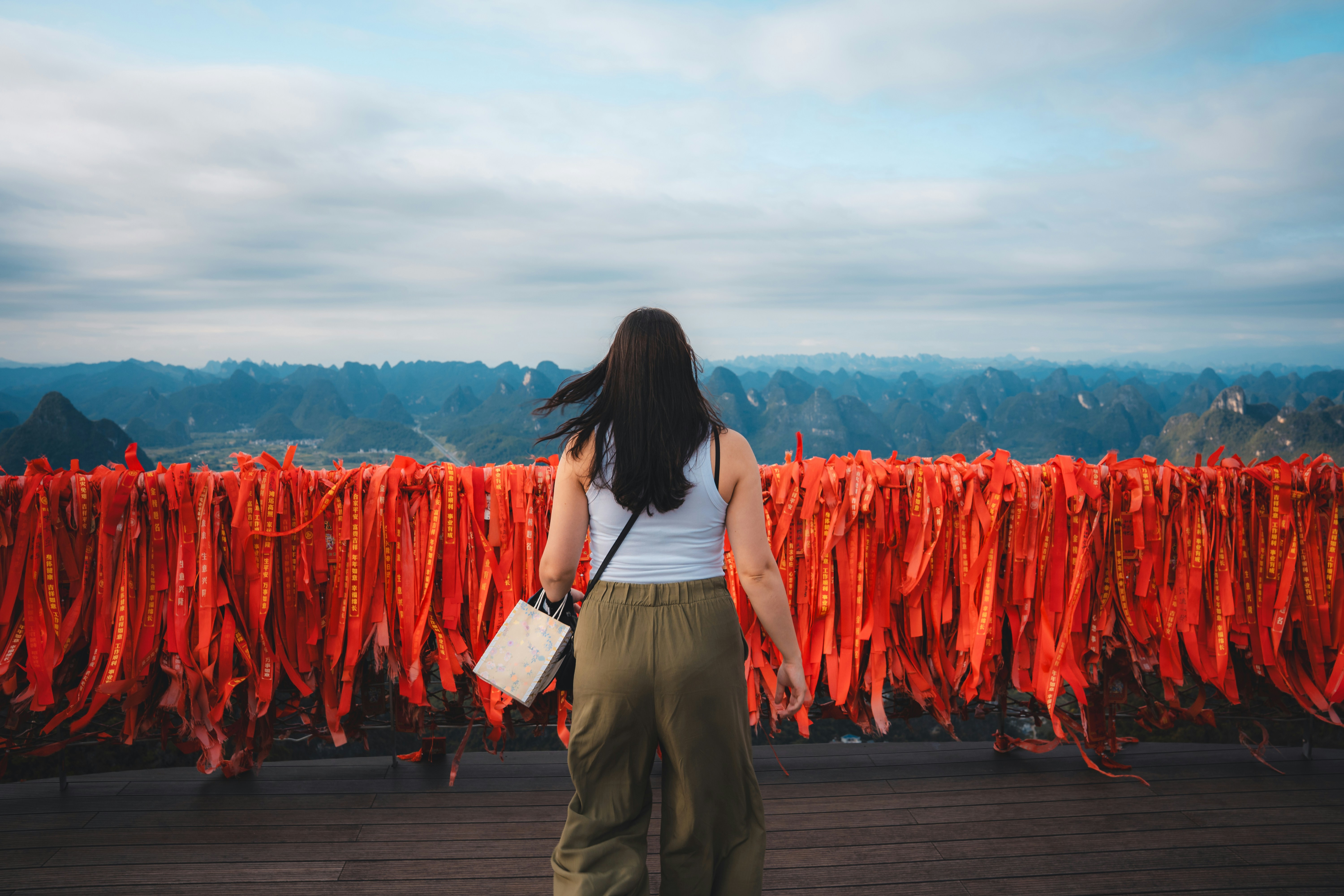 Woman in white top and olive pants foregrounds a sea of red ribbons against a distant mountain panorama