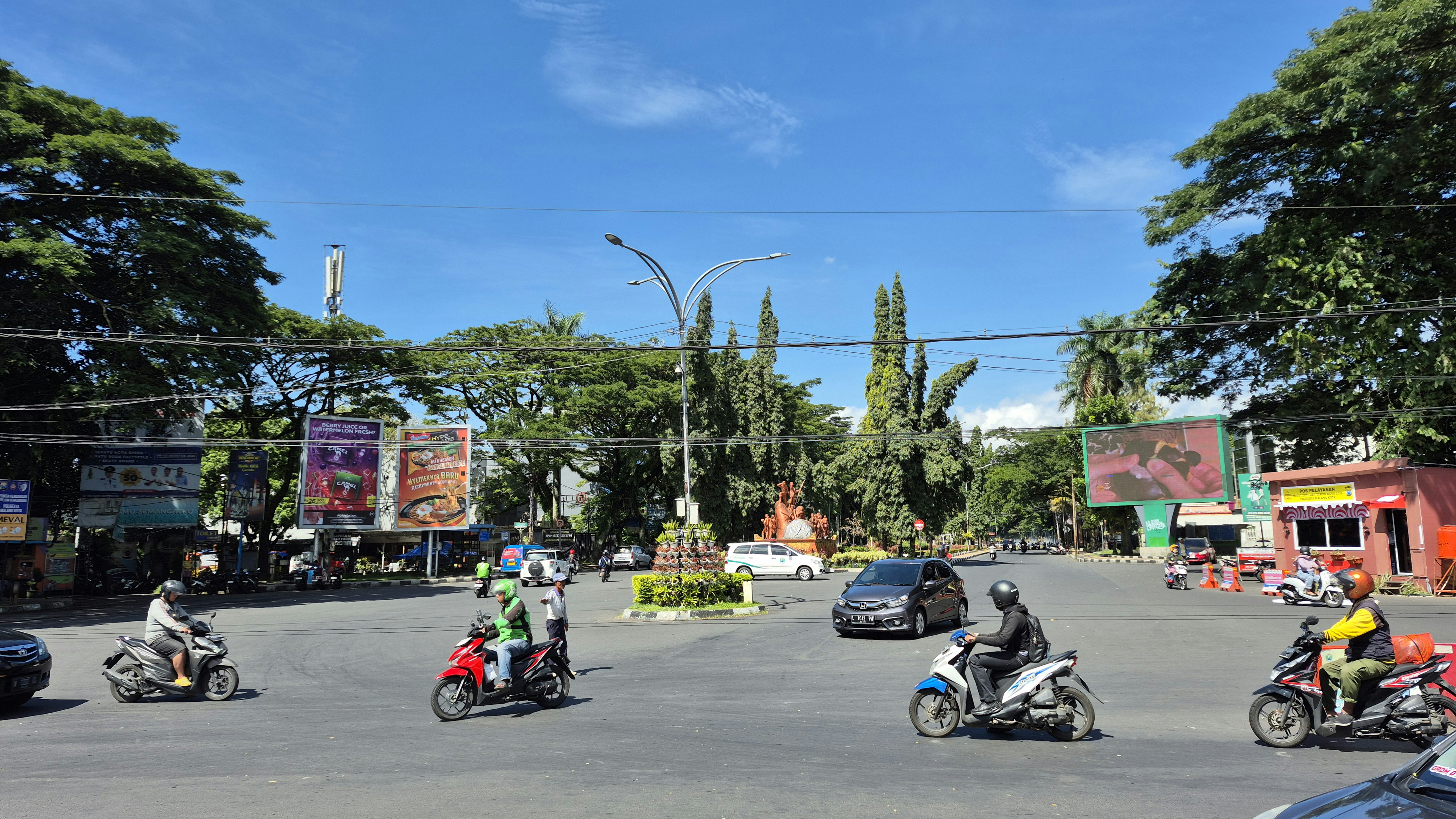 Motorcycles and cars navigate a busy intersection with trees.