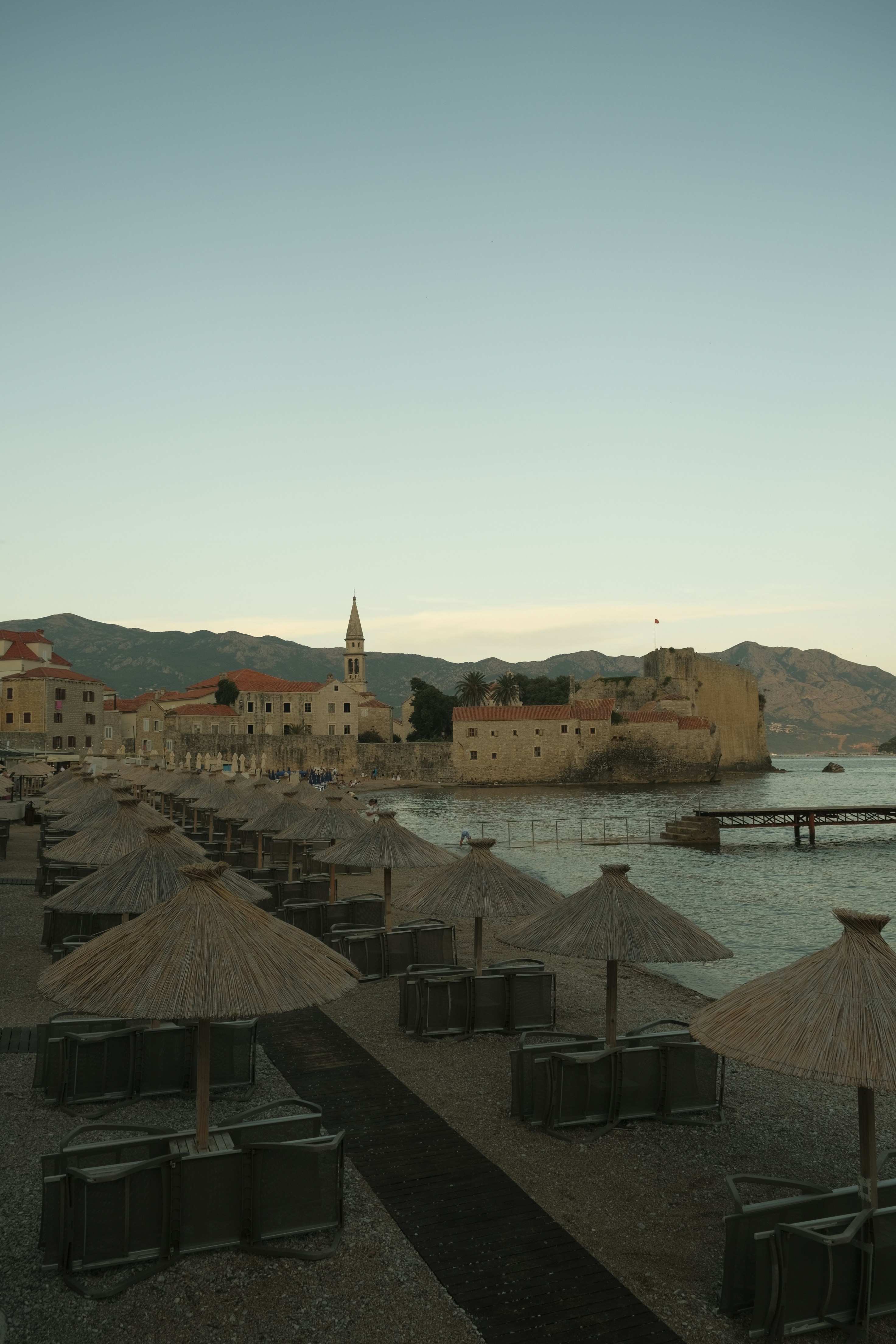 Sunlit Croatian shore: straw parasols align toward a distant fortress
