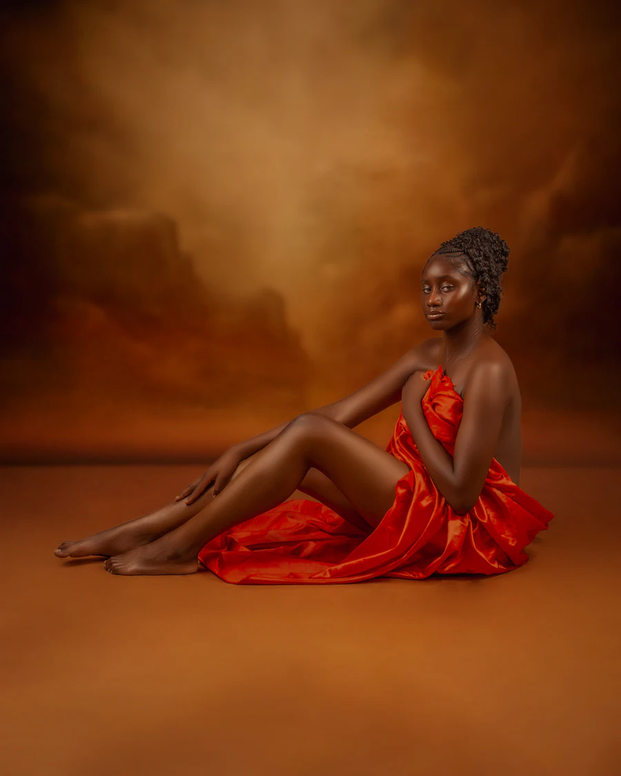 A young woman sits gracefully on a warm amber backdrop, wrapped in a vivid red fabric that pools around her. The portrait emphasizes natural poise and smooth skin tones against a soft, gradient background.