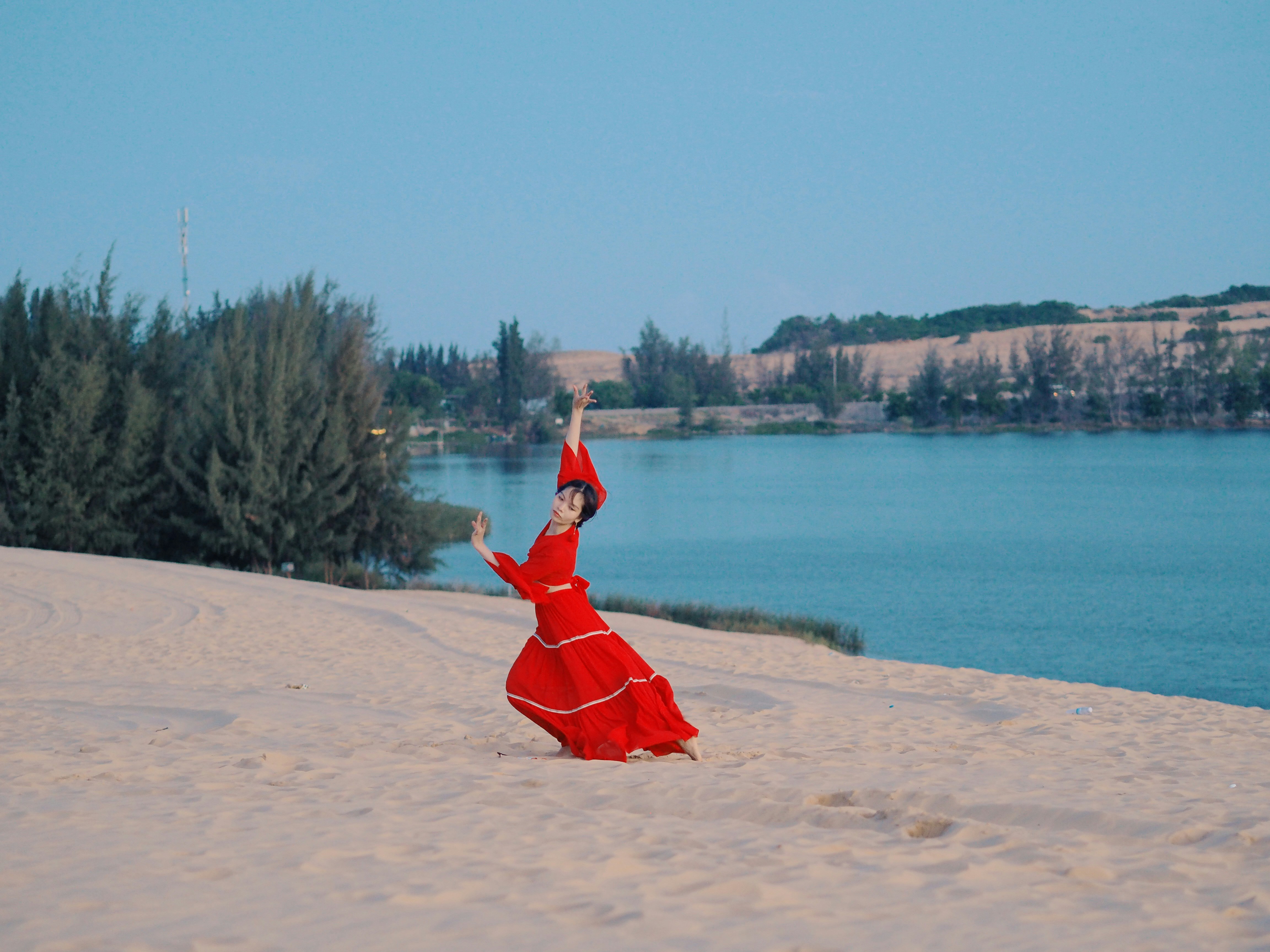Red-dressed dancer on sunlit dunes by a tranquil blue inlet
