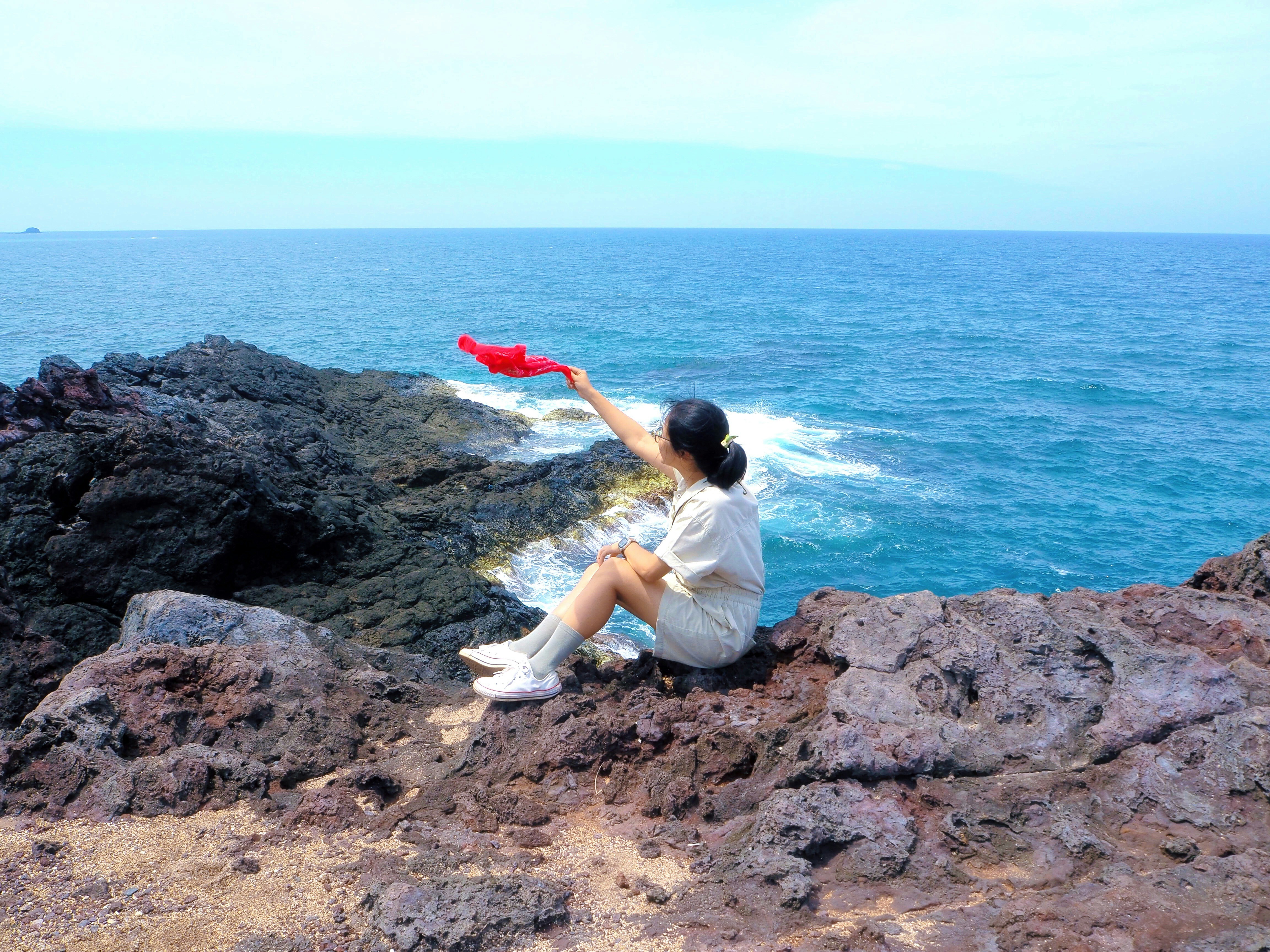 Seaside Moment: A lone figure on volcanic rocks releasing a red scarf toward the sea