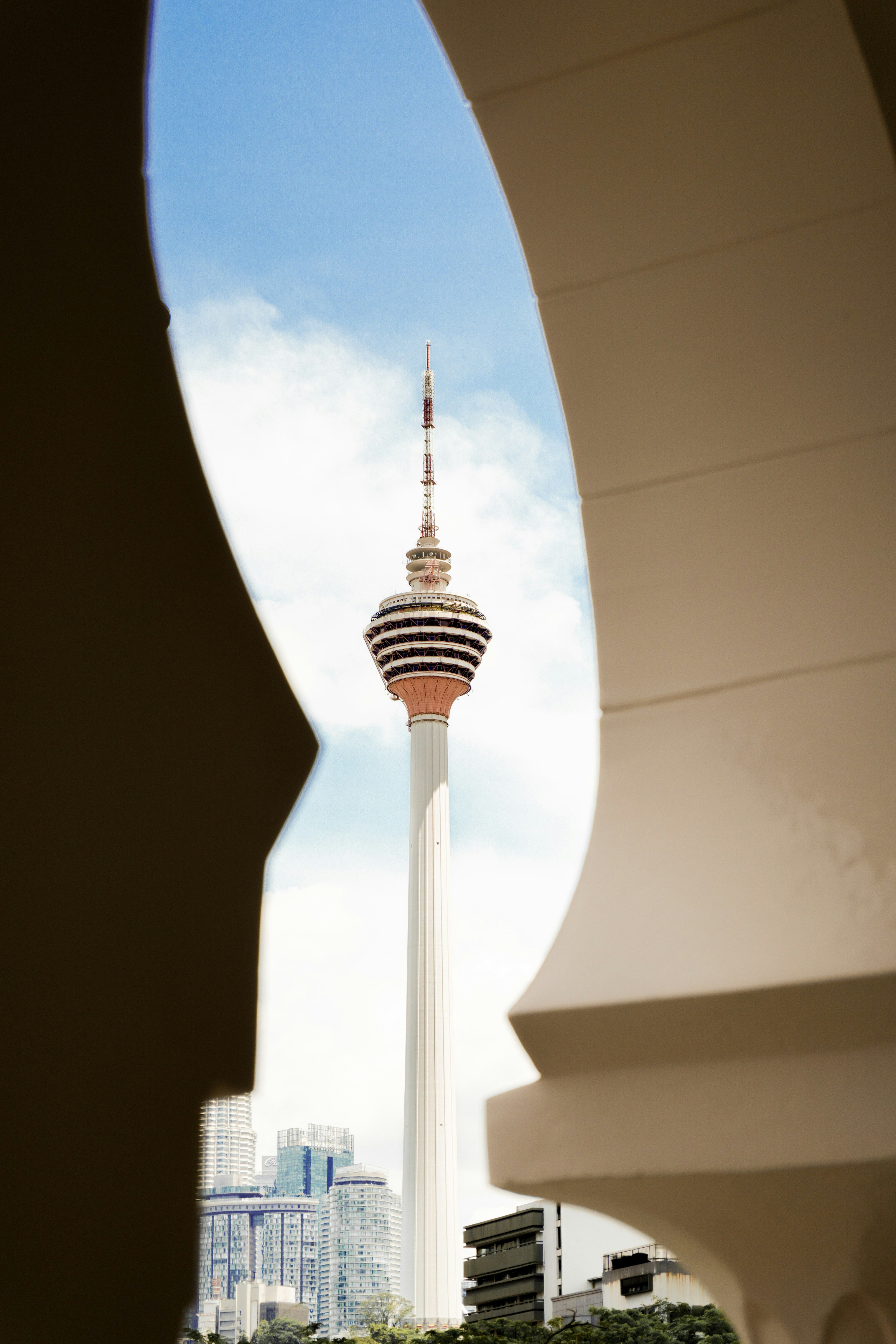 Towering Kuala Lumpur's communications mast framed by architectural arches in foreground, with cityscape below. A minimalist architectural study emphasizing vertical lines and negative space.