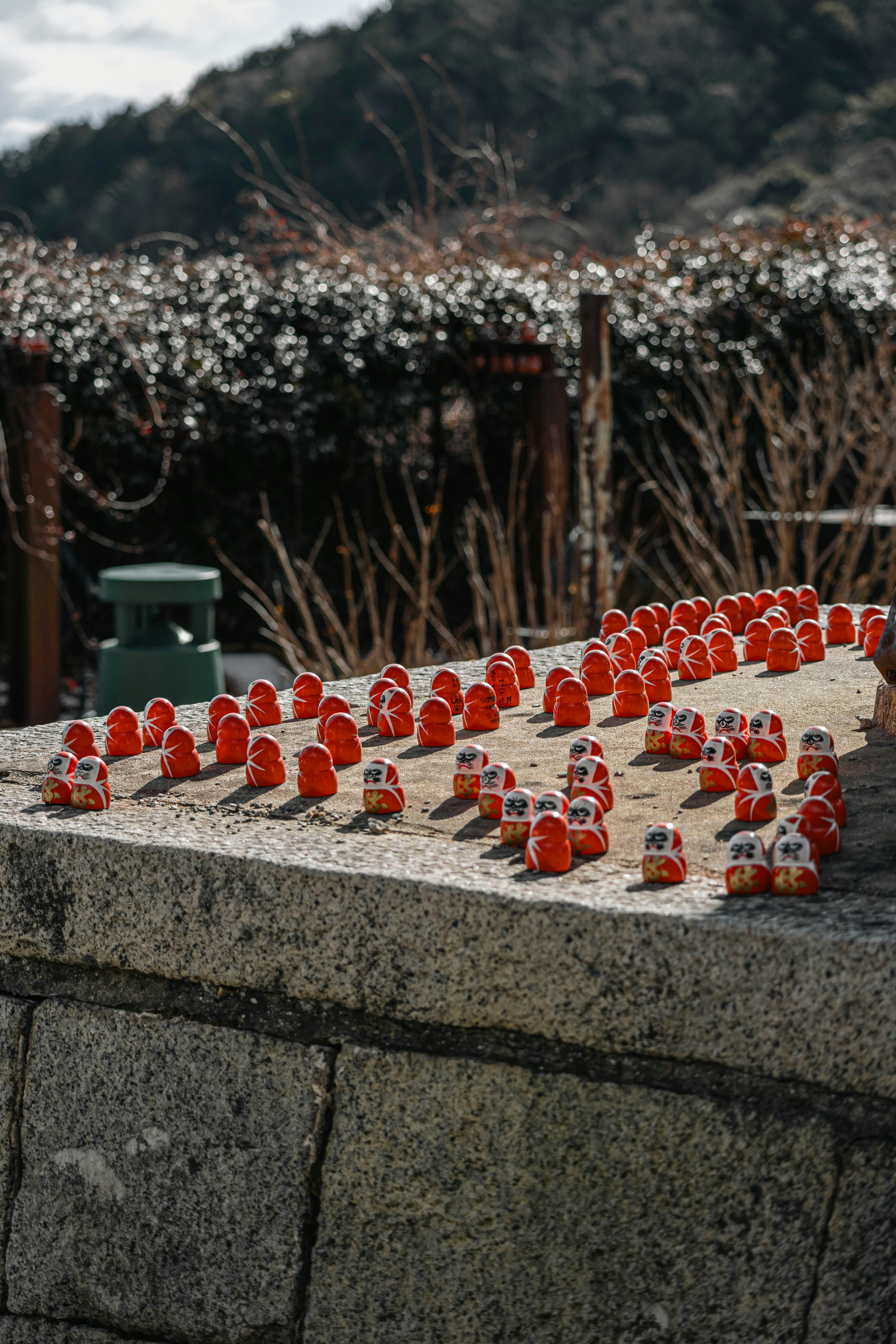 Row of red Daruma dolls on a stone ledge with a blurred garden backdrop
