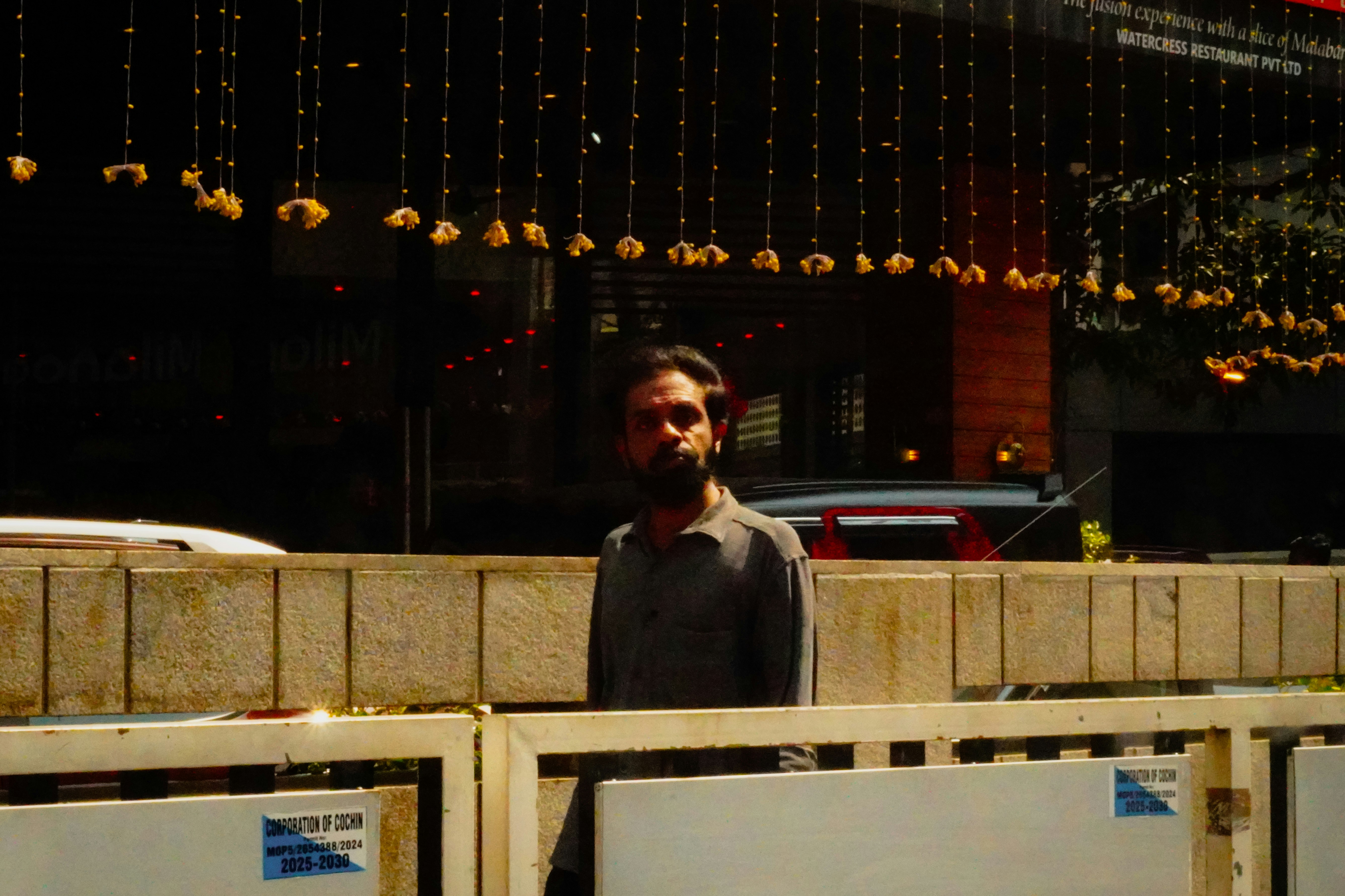 Man in a gray shirt stands behind a concrete barrier under string lights