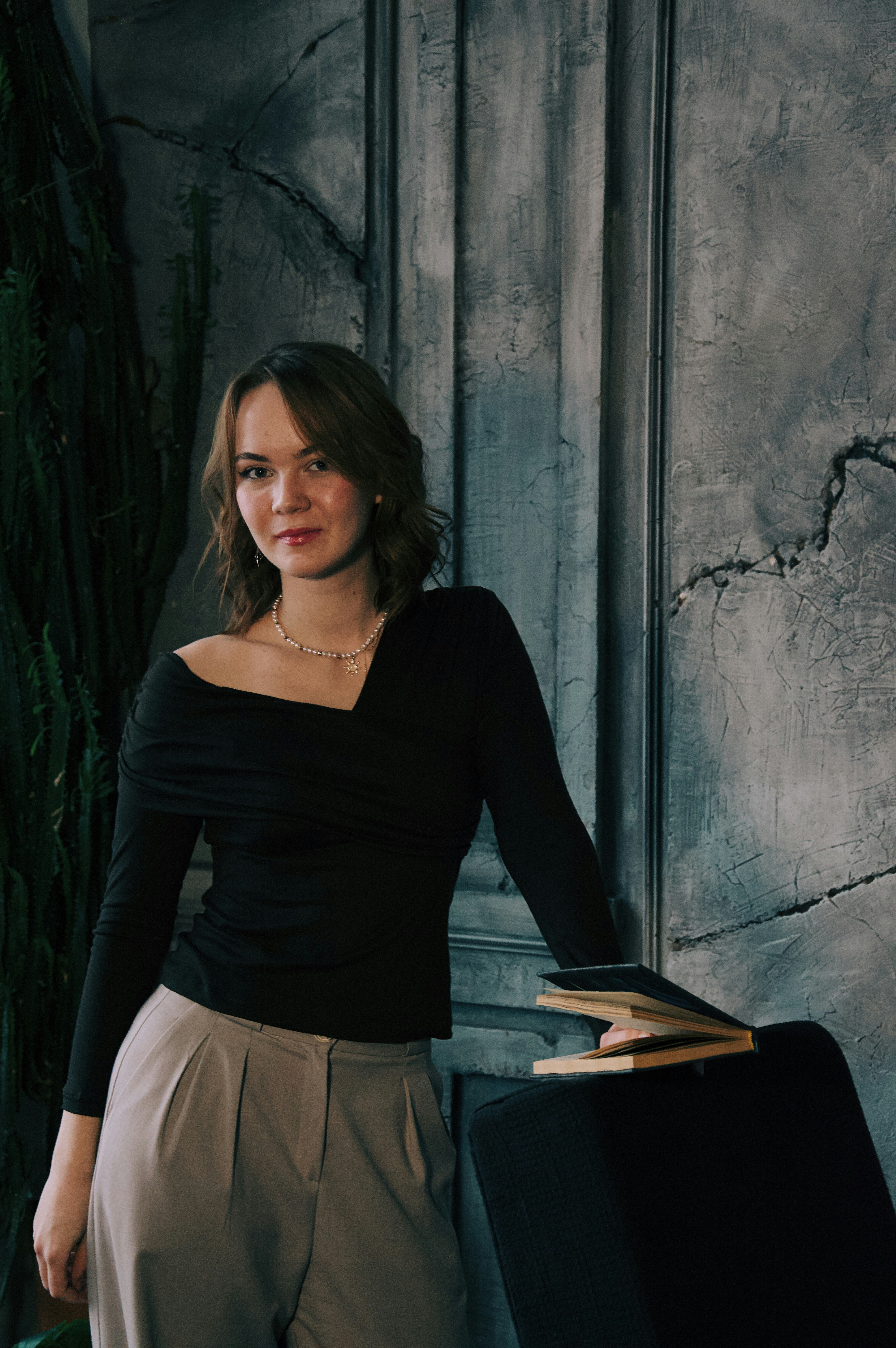 Elegant Poise: A woman in a black off-shoulder top leans beside a chair with a pair of books.