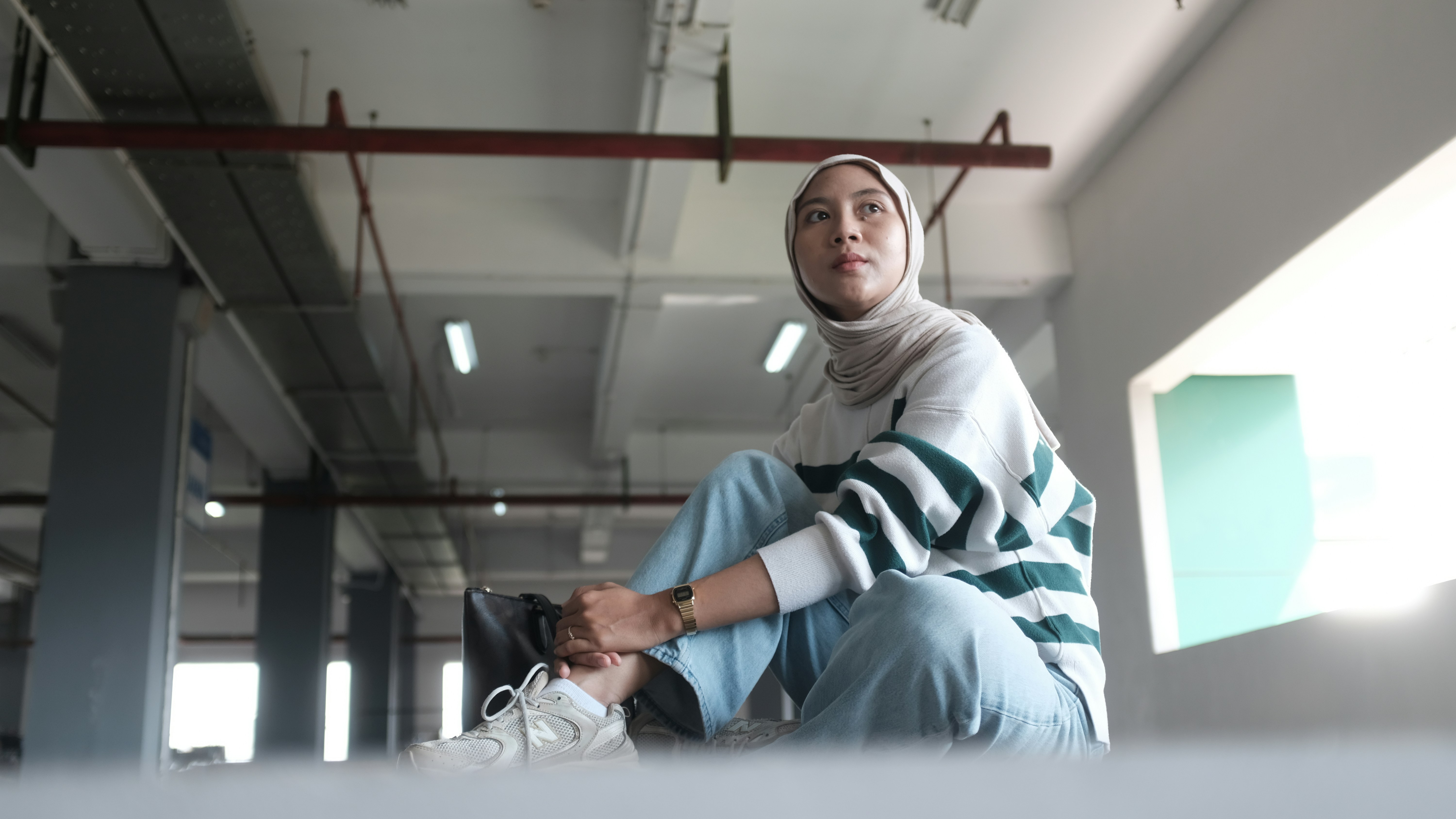 Hungry Lounger: A hijab-clad traveler rests on a concrete ledge in an industrial-lit space