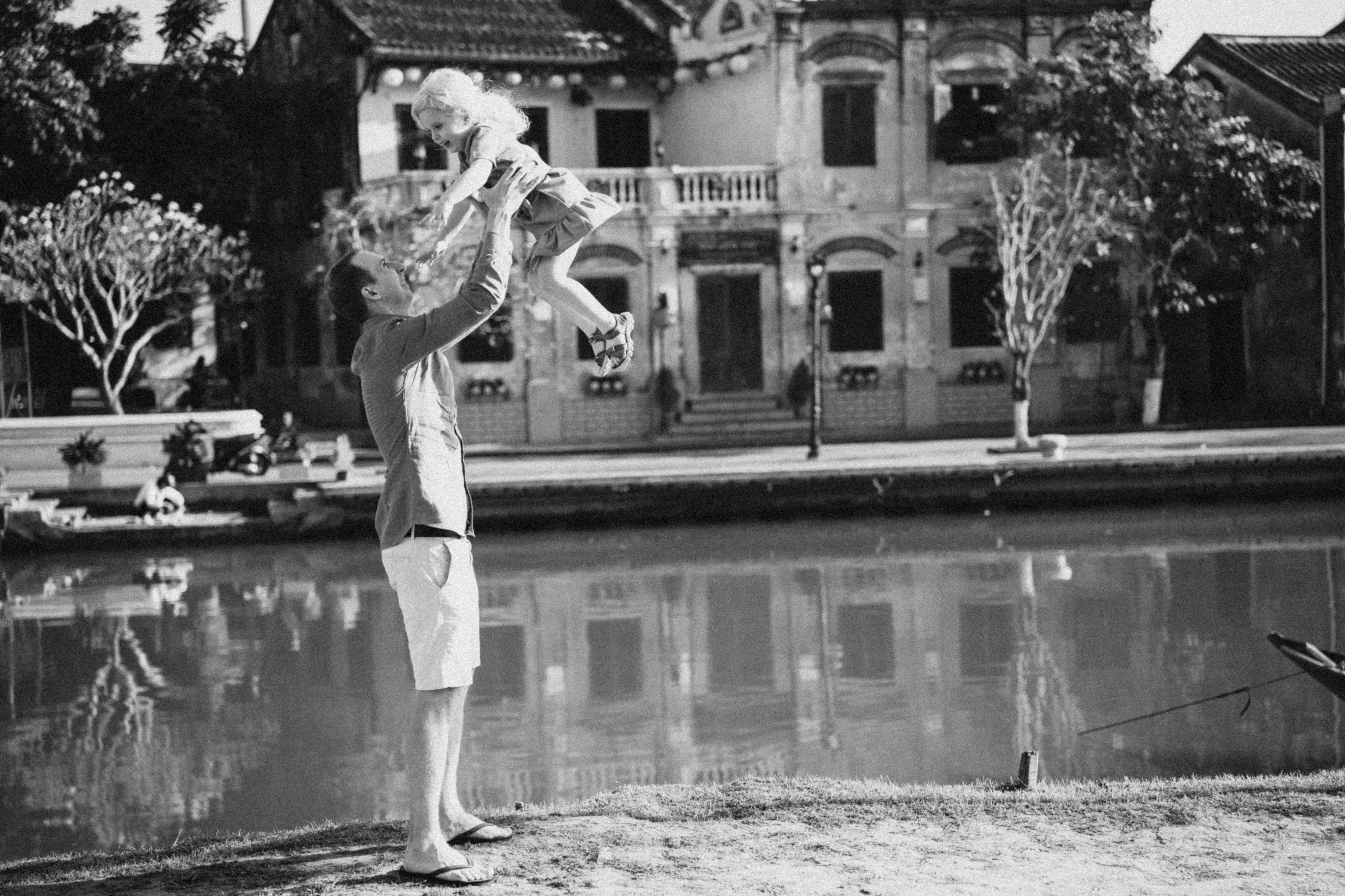 Lift and Laugh: Father and Child in a Sunlit Riverside Courtyard