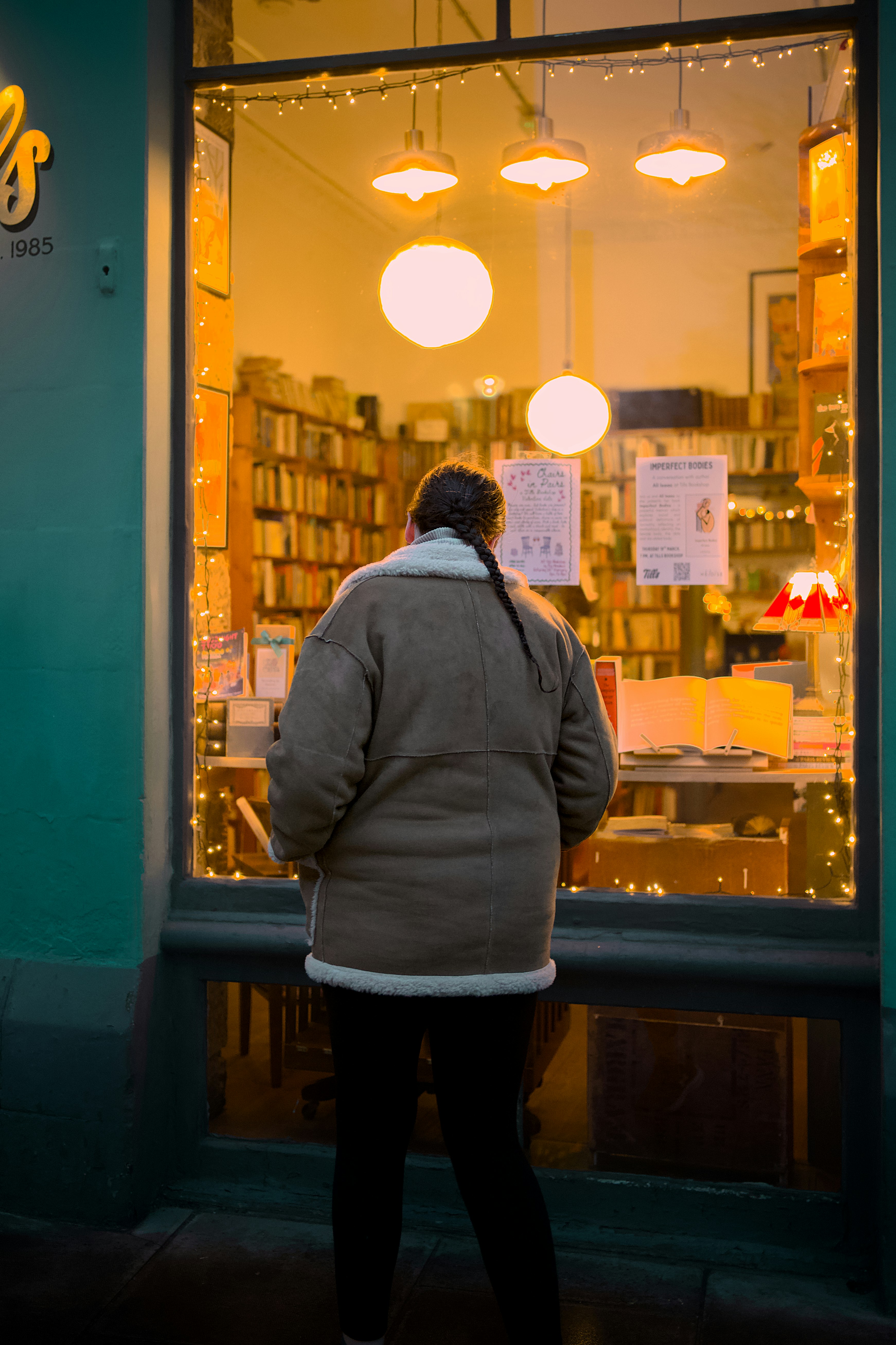 Warm storefront glow: a lone figure absorbed in a bookshop night