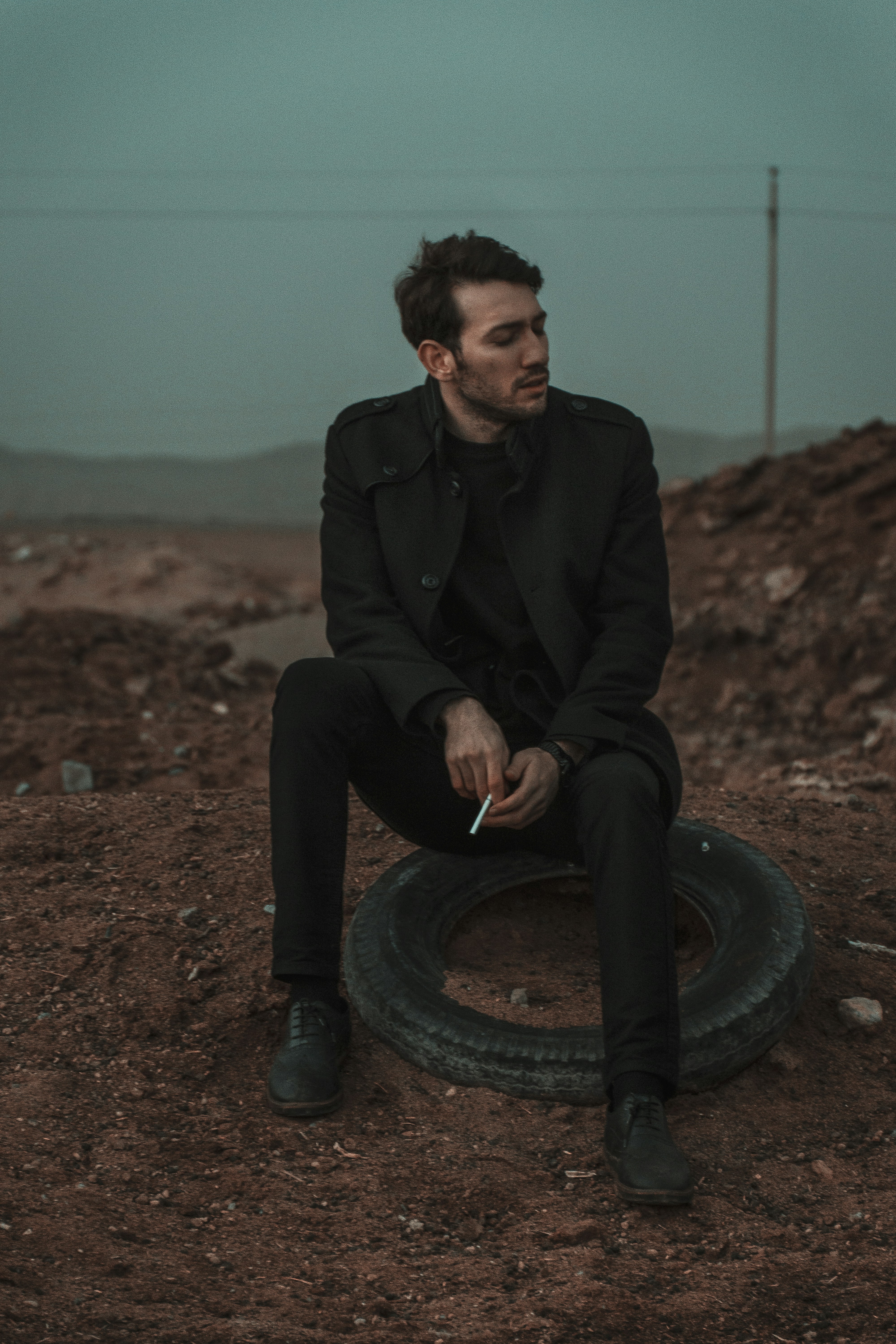 Deserted Talk: A man in dark coat sits on an old tire, cigarette in hand, under a somber sky