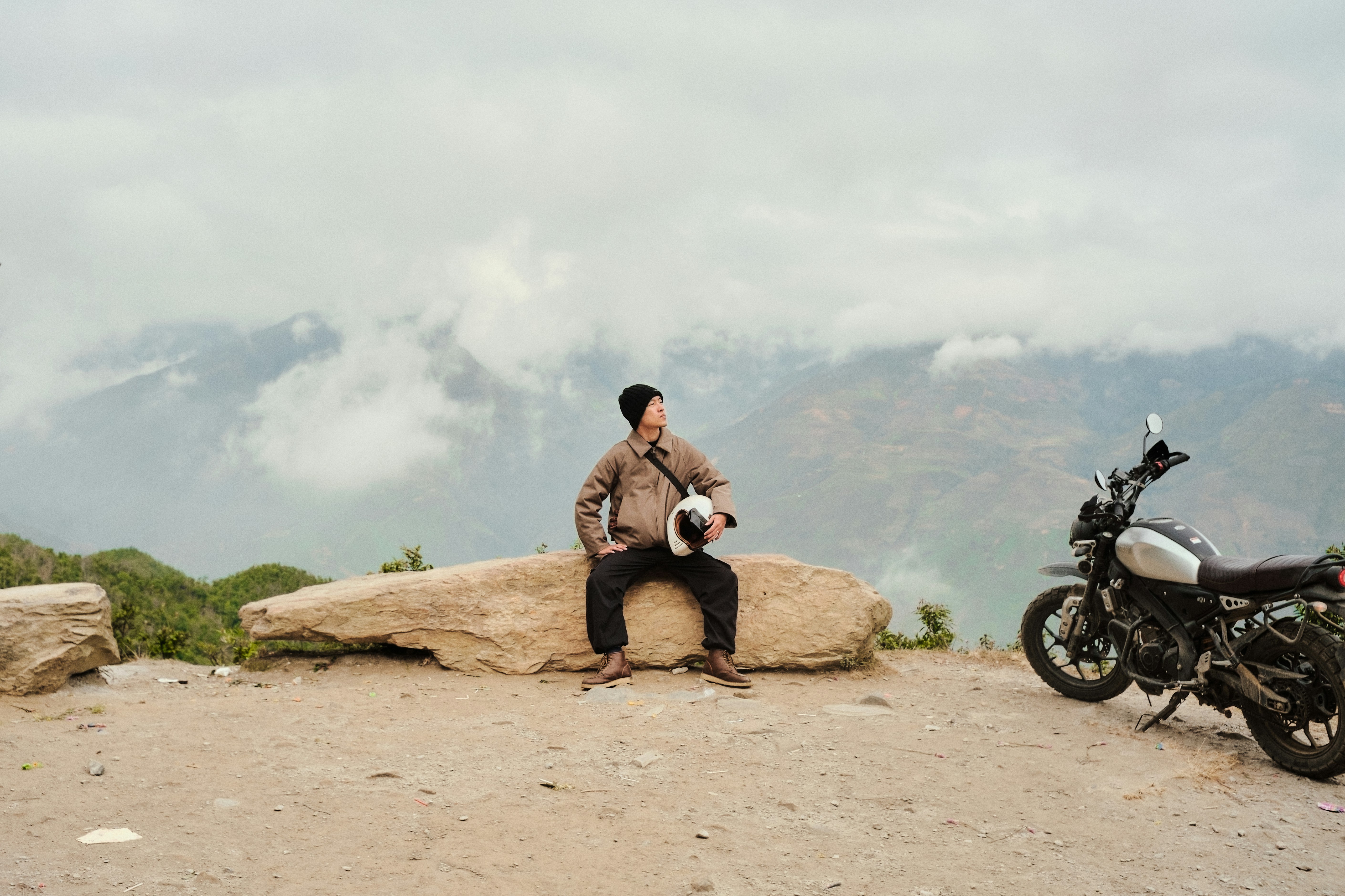 A solitary man sits on a rock with a helmet in hand, overlooking a distant landscape; a motorcycle rests to the right on a dusty overlook.
