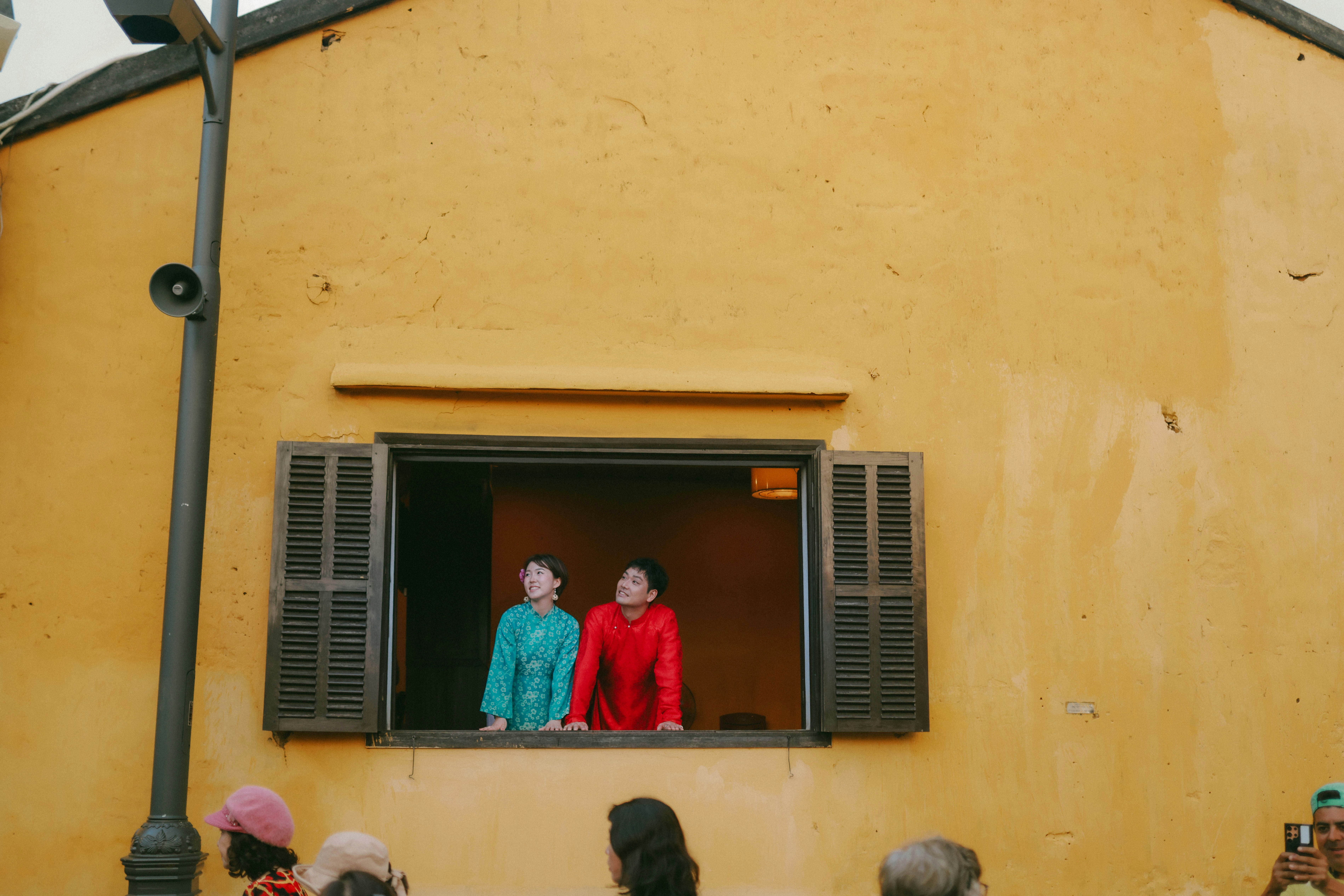 Two figures at a sunlit window watching over a yellow courtyard
