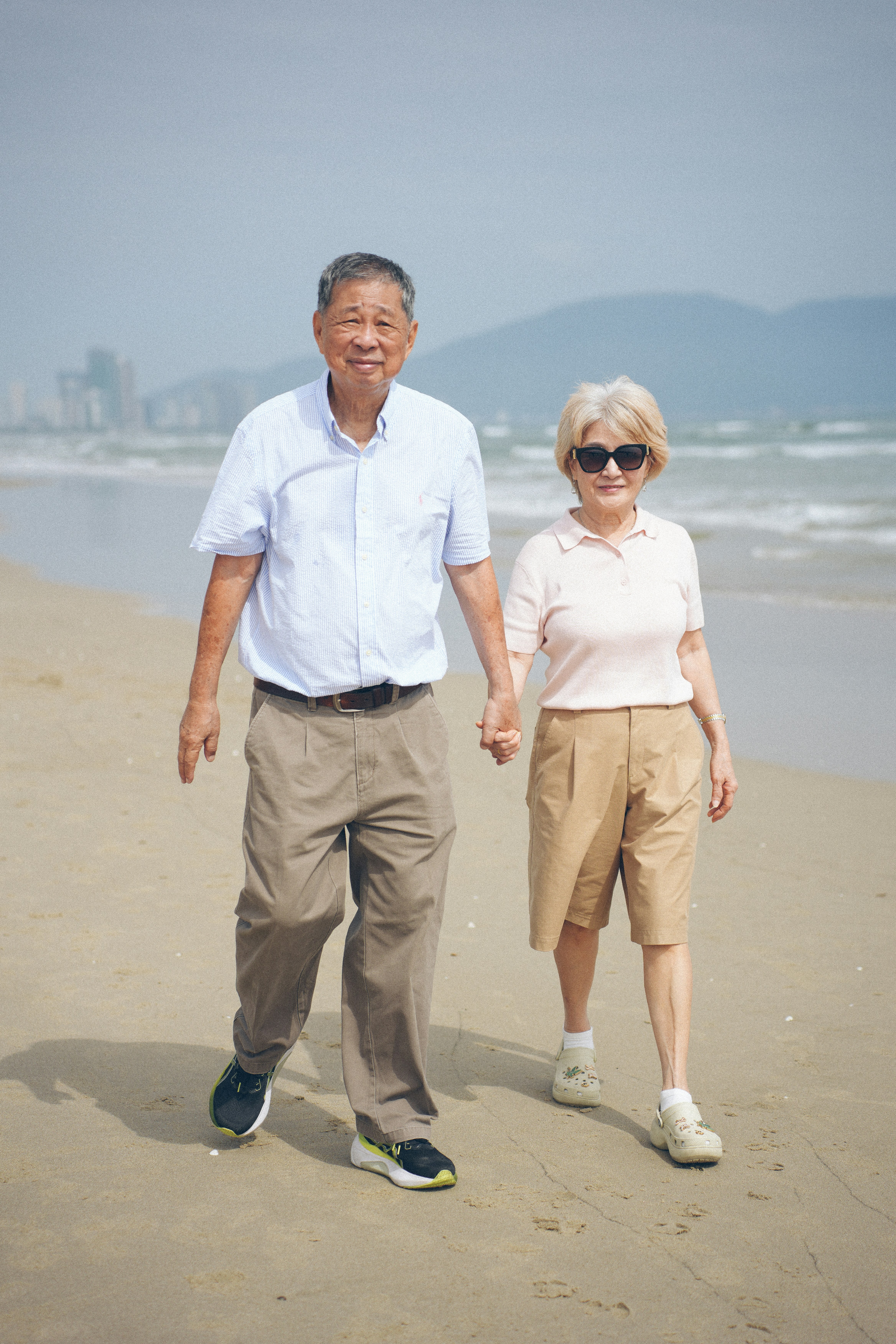 Elderly couple holding hands walking on a sandy beach.