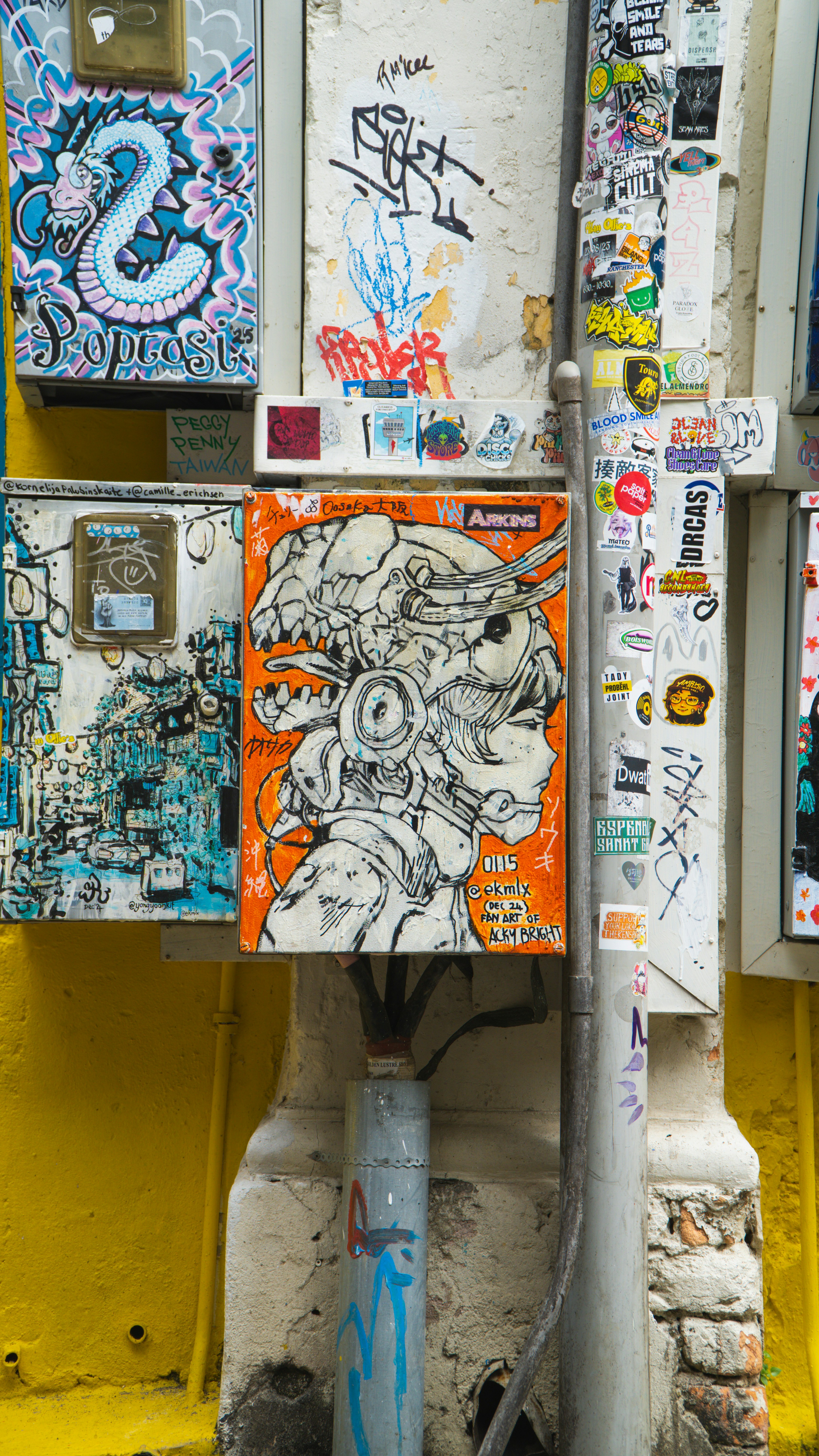 Street Art Mosaic: Orange Creature Painting Anchors a Wall of Stickered Panels