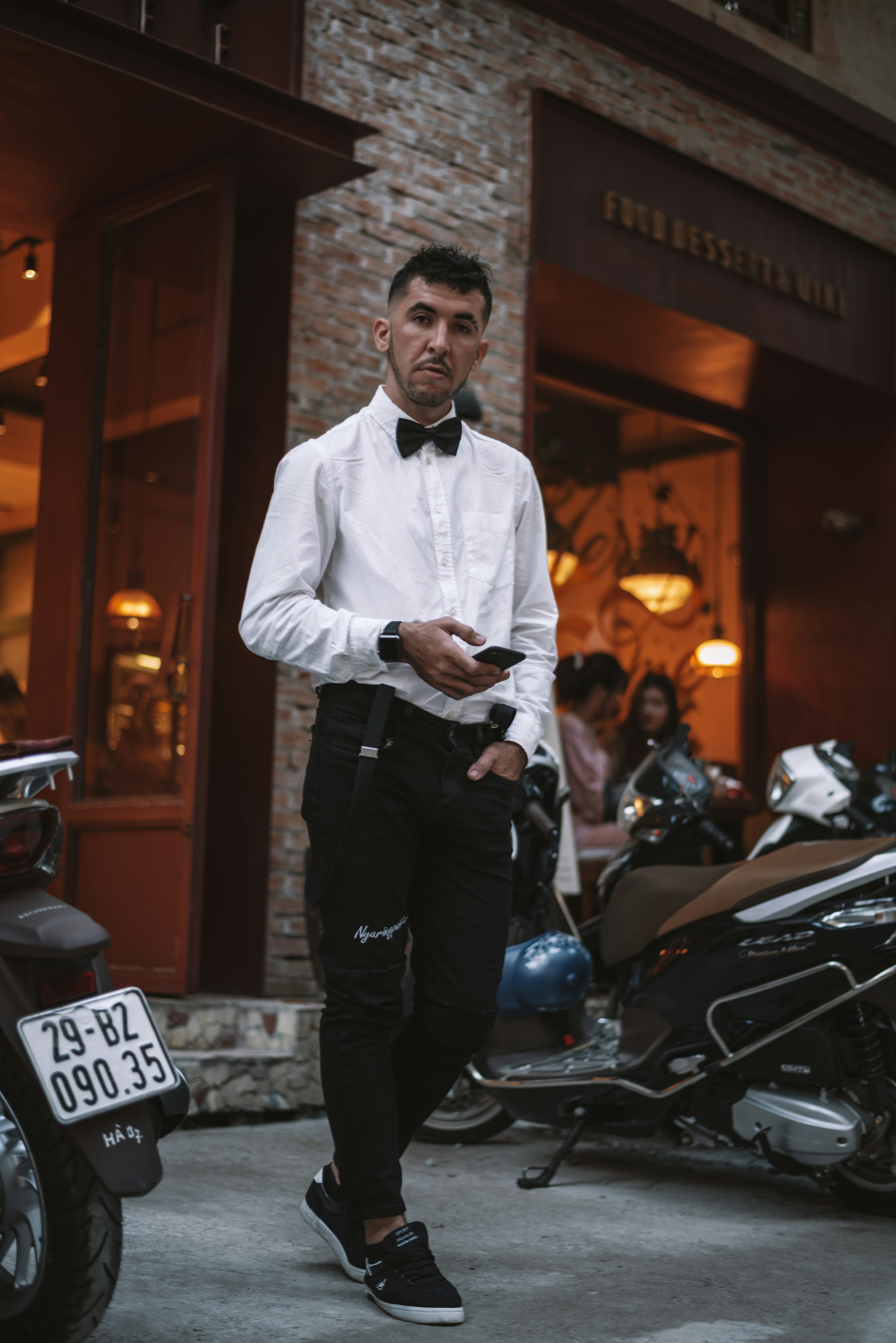 Gentleman in White Shirt with Bow Tie Holds Phone Outside a Brick-Wuild Café
