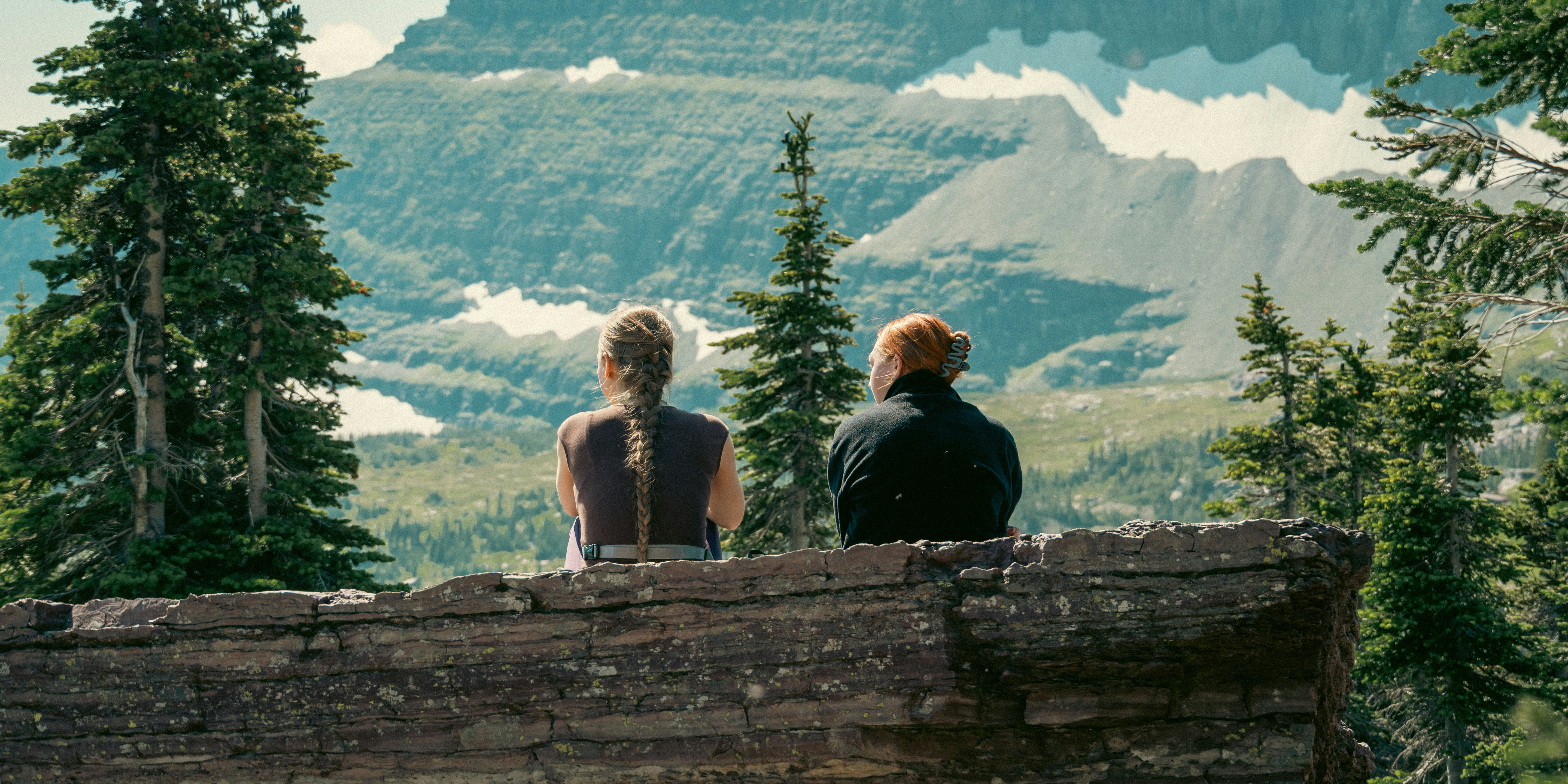 Two silhouettes on a rocky ledge overlook a mountain valley