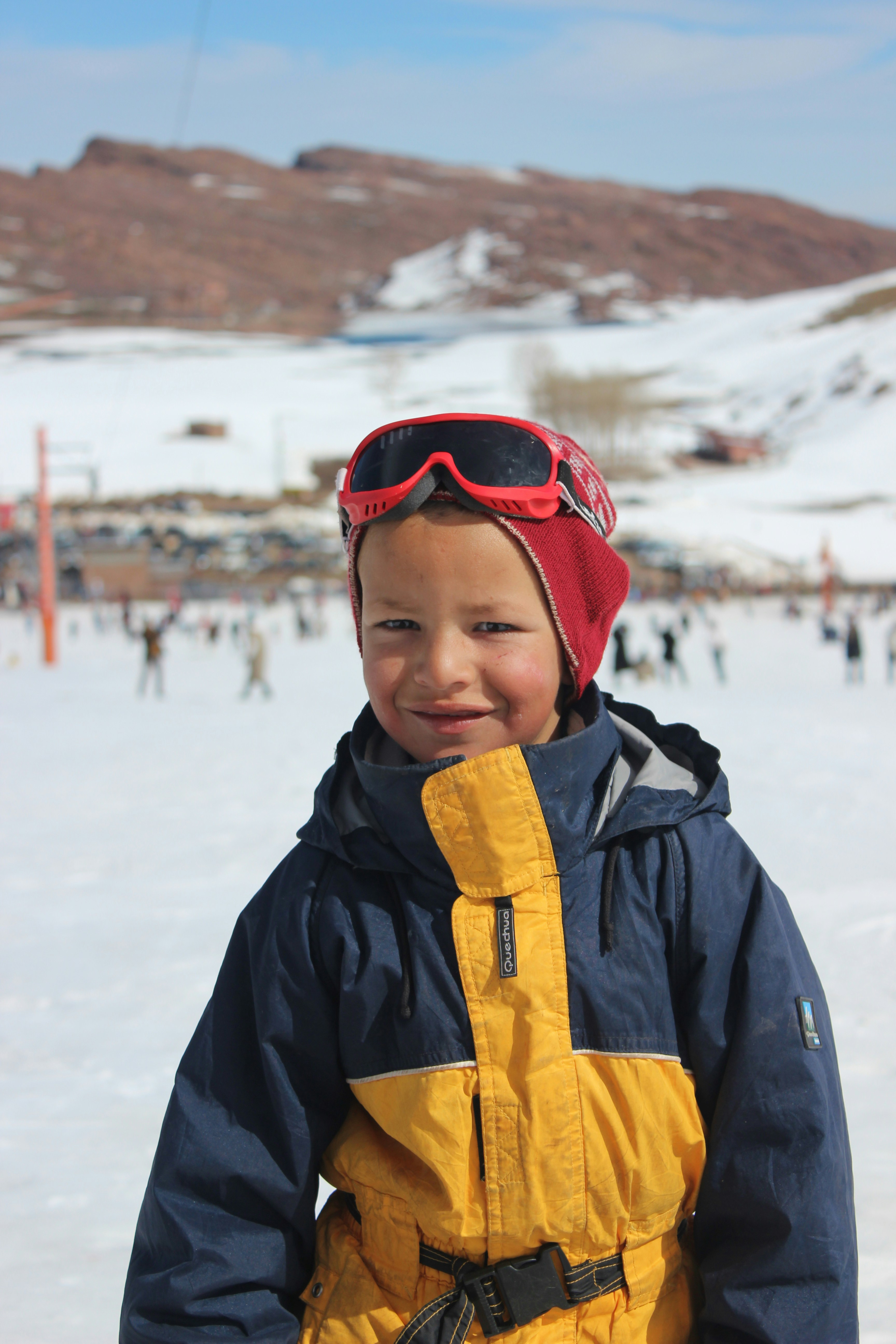 Child in bright yellow-and-navy snowsuit with red goggles in a sunlit alpine scene
