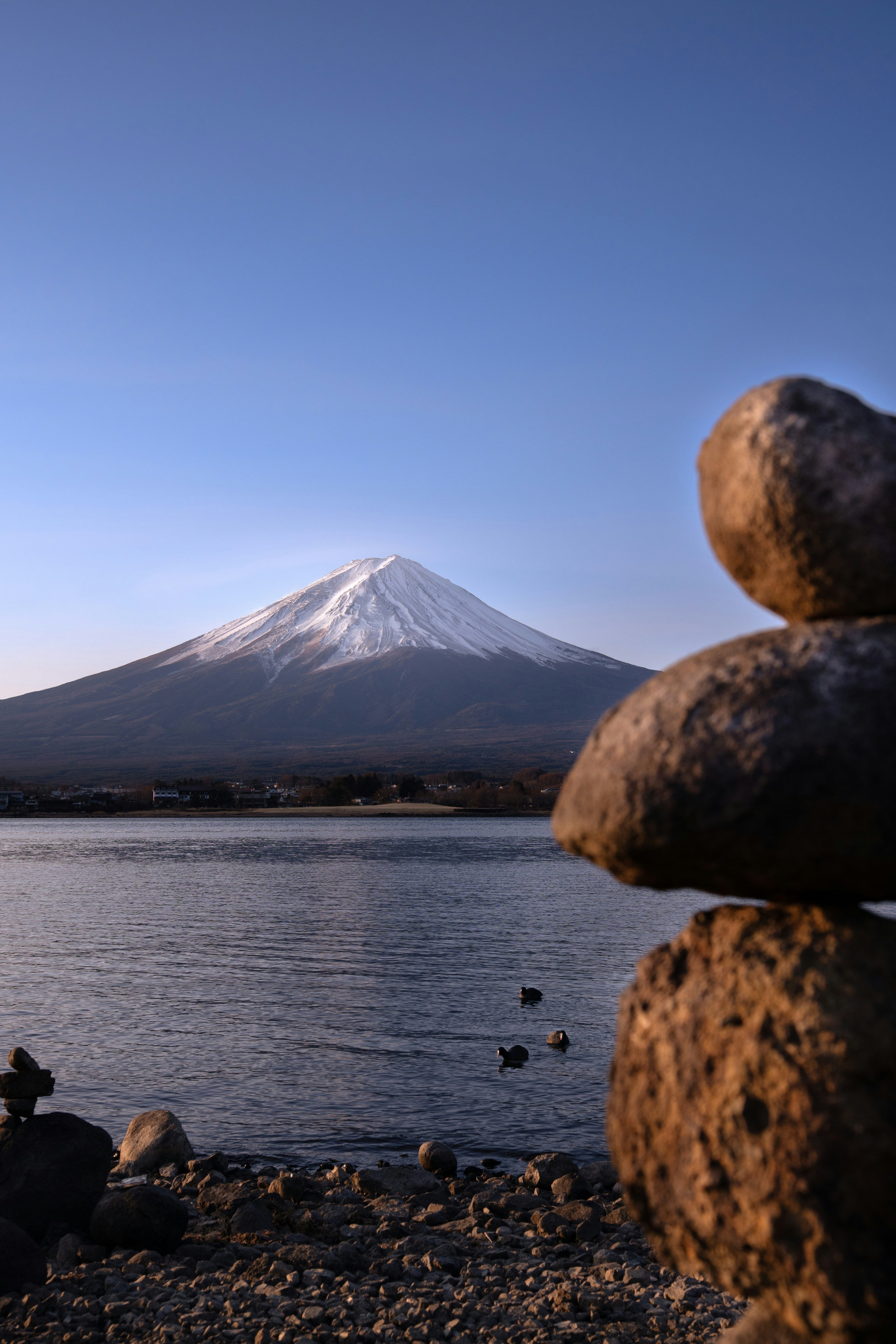 Snow-capped Fuji over a tranquil lakeshore with stacked stones in foreground