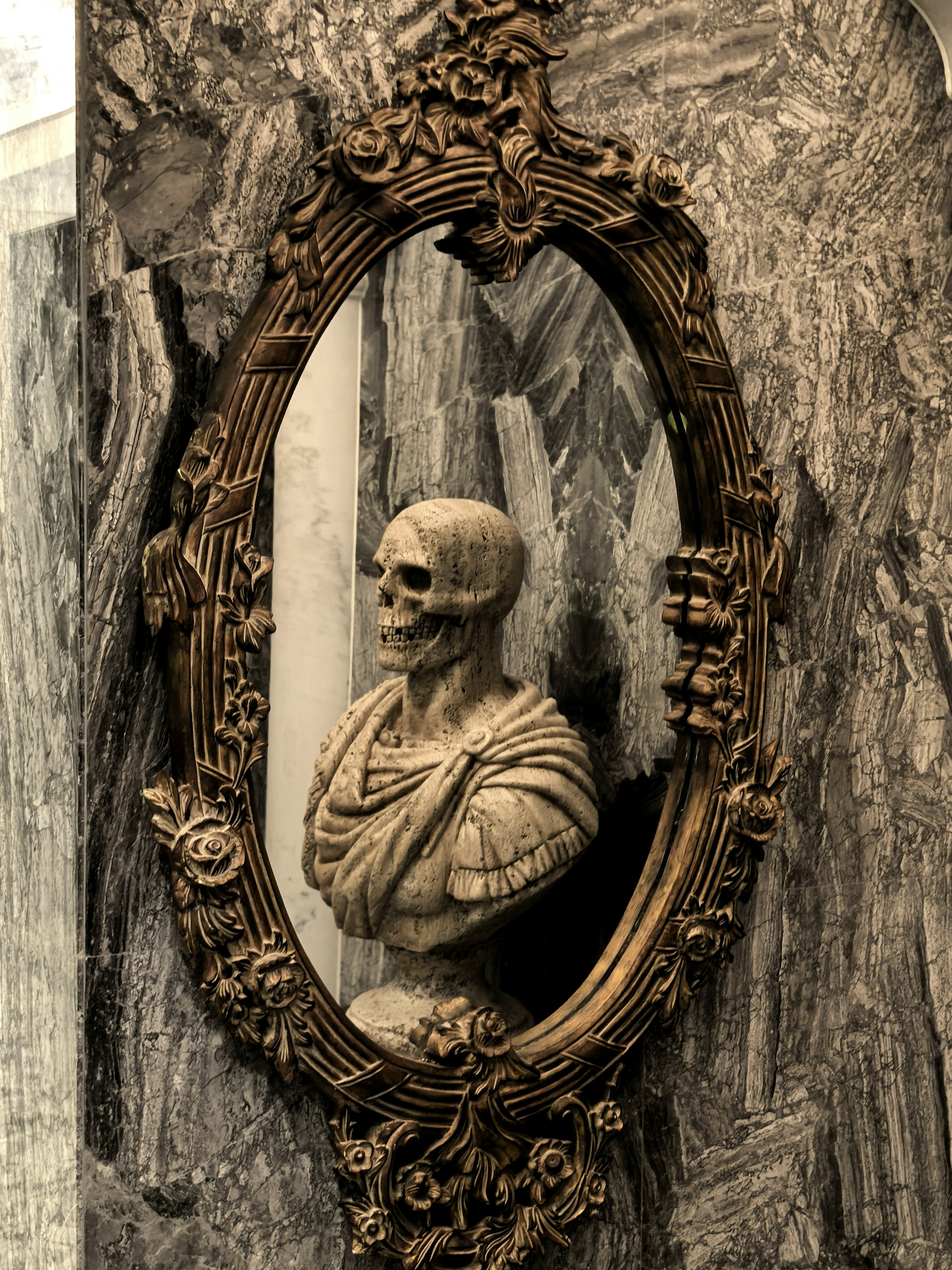 Ornate mirror reflects a marble bust of a skeleton.
