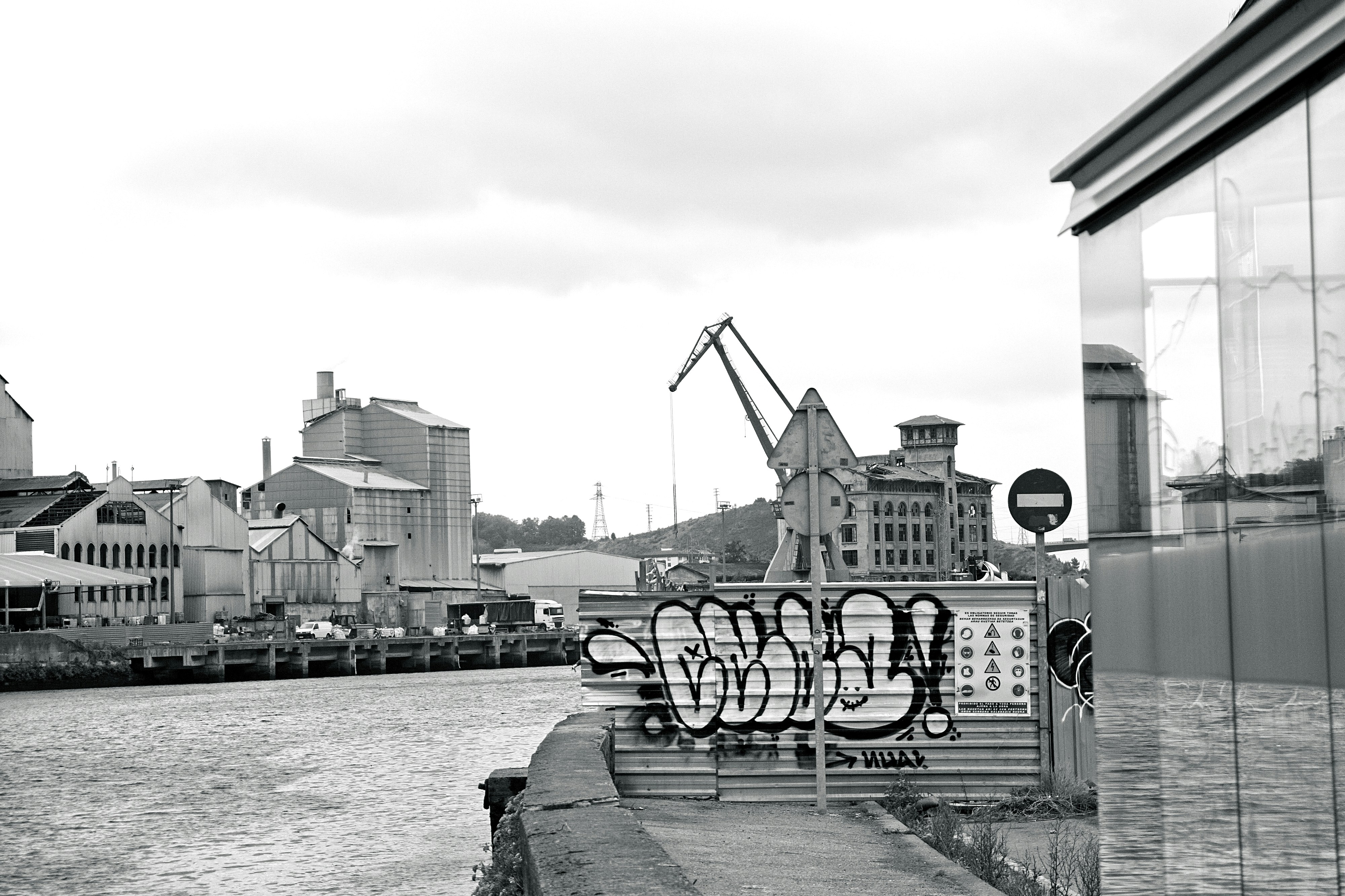 A grayscale riverside industrial area with graffiti on a metal barrier along the quay, extending toward a crane and factory buildings in the distance.