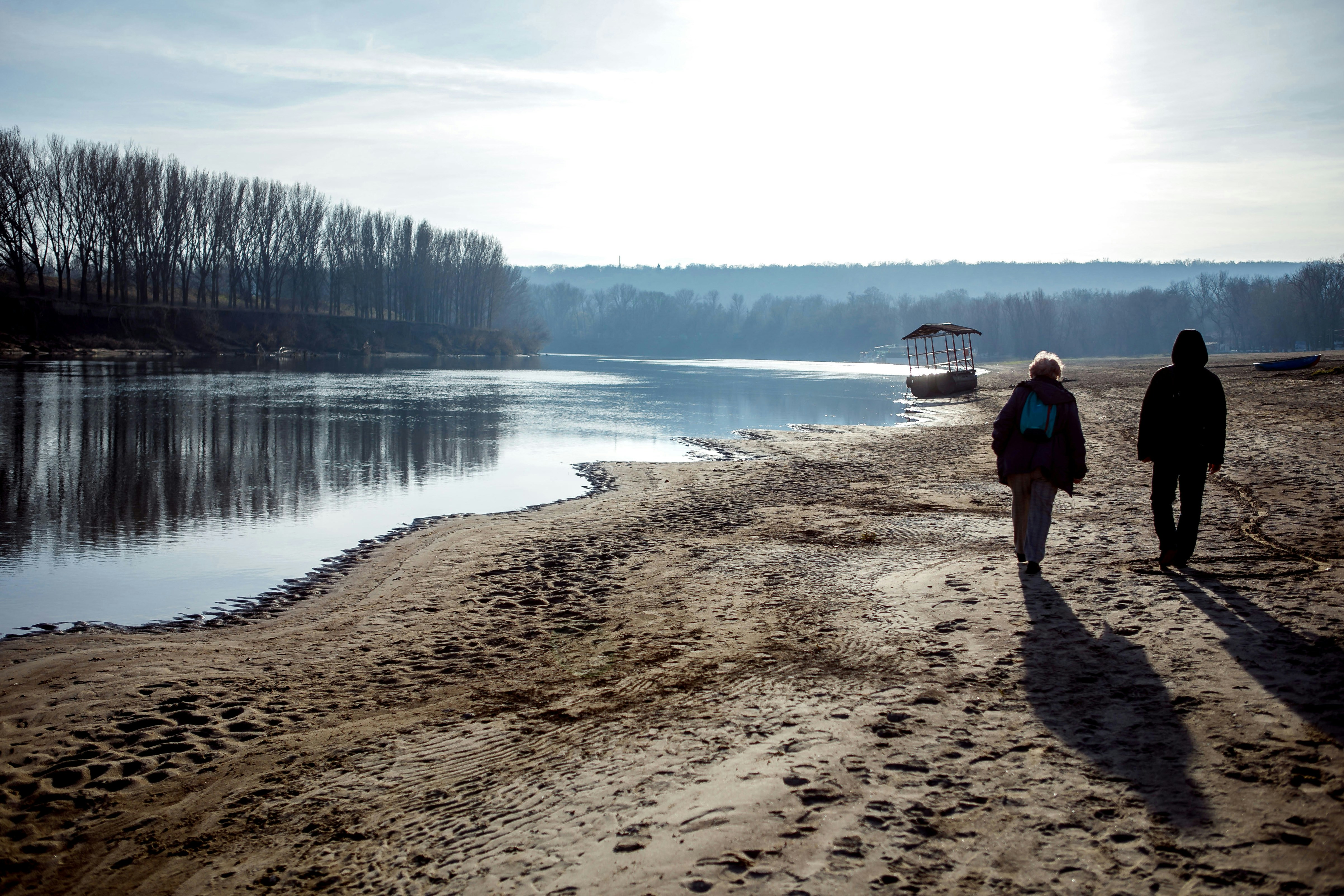 Two walkers at the riverside trailing into the light, near the docked boat