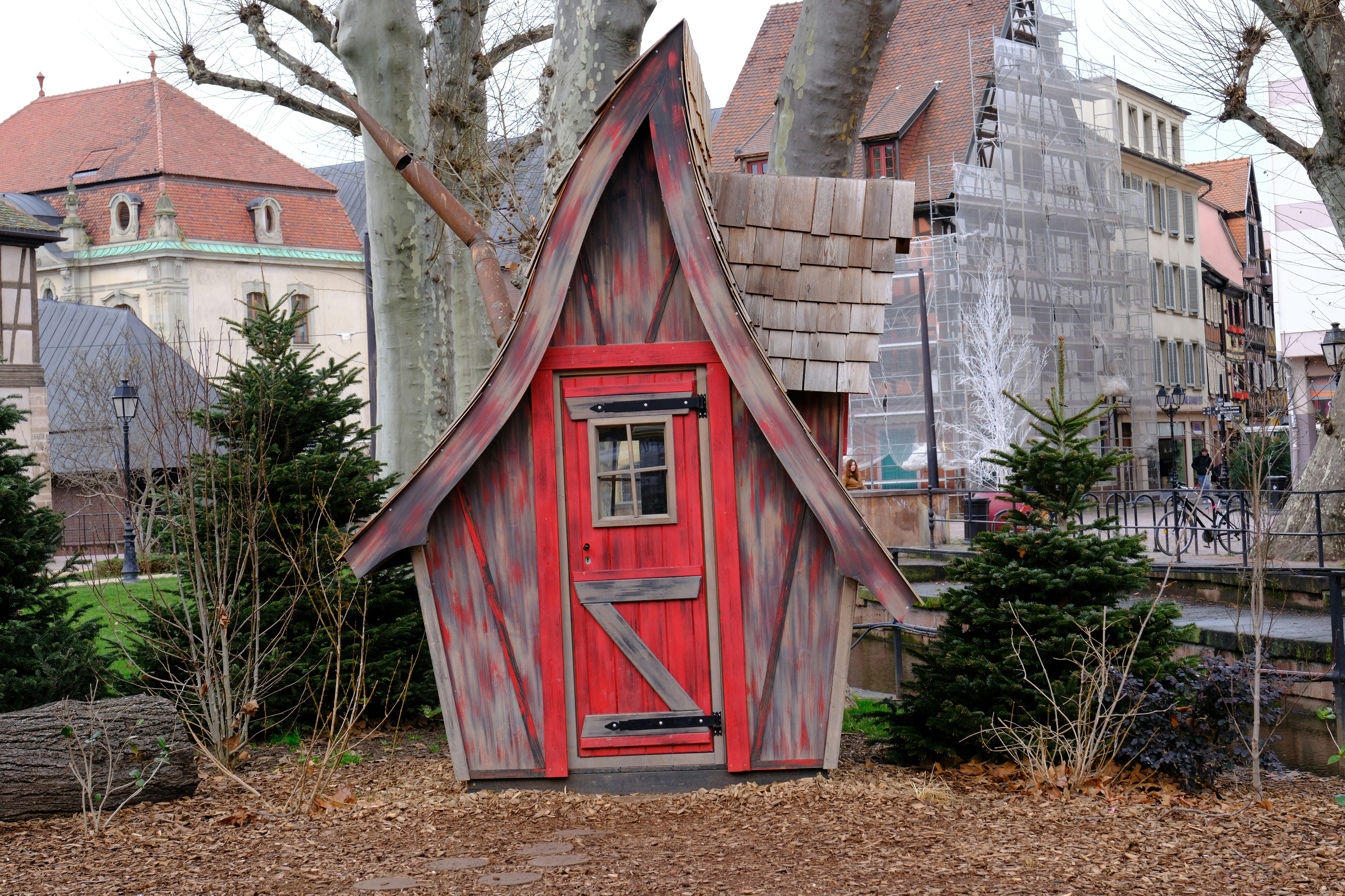 Vivid red wooden hut sits in a city square, surrounded by small evergreens and bare ground; modern buildings and a scaffolded facade provide a contrasting backdrop.