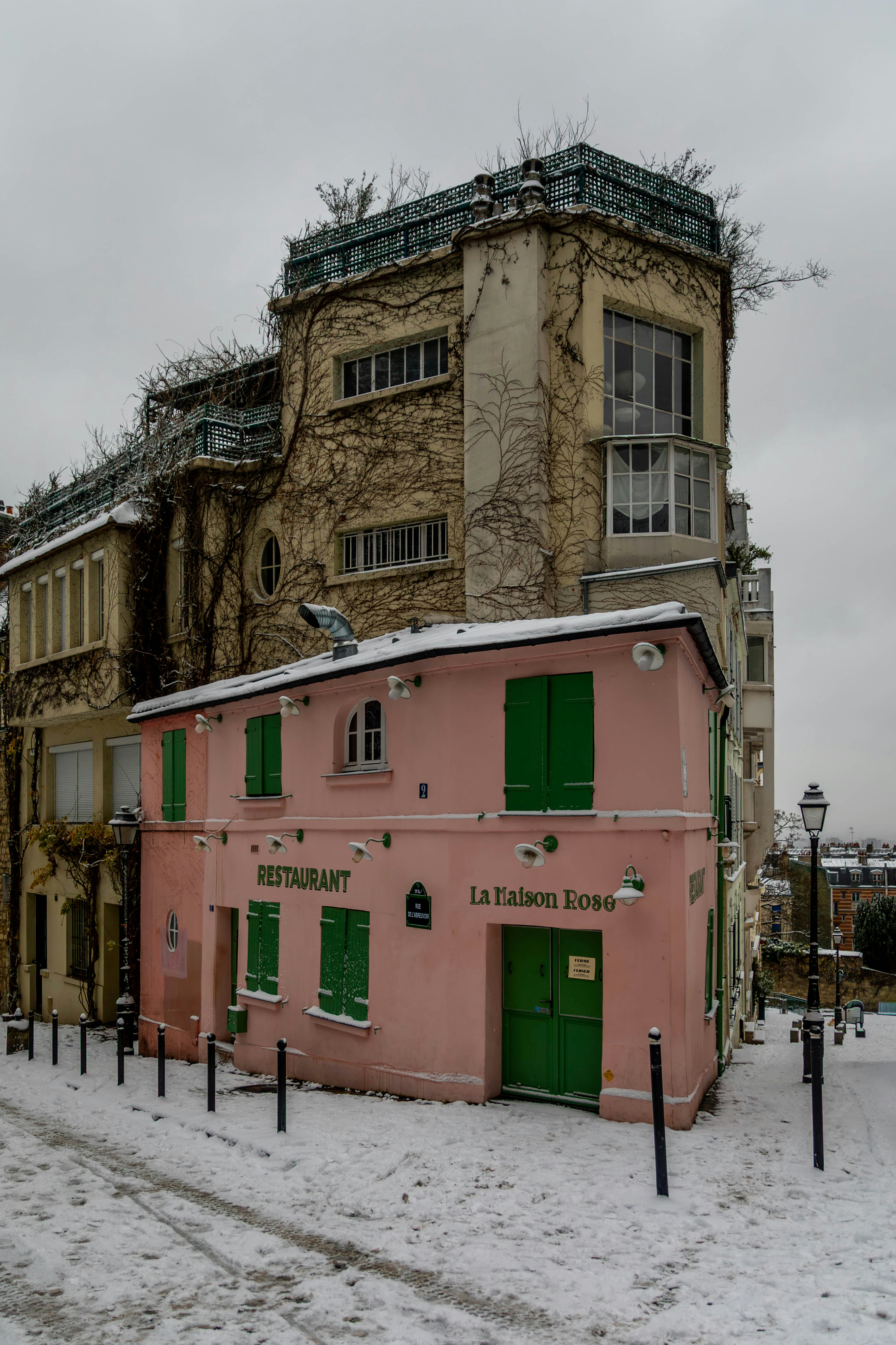 Snow blankets a pink restaurant building with vivid green shutters, framed by a taller limestone structure; overcast sky and vine-wrapped walls create a moody, wintry street scene.