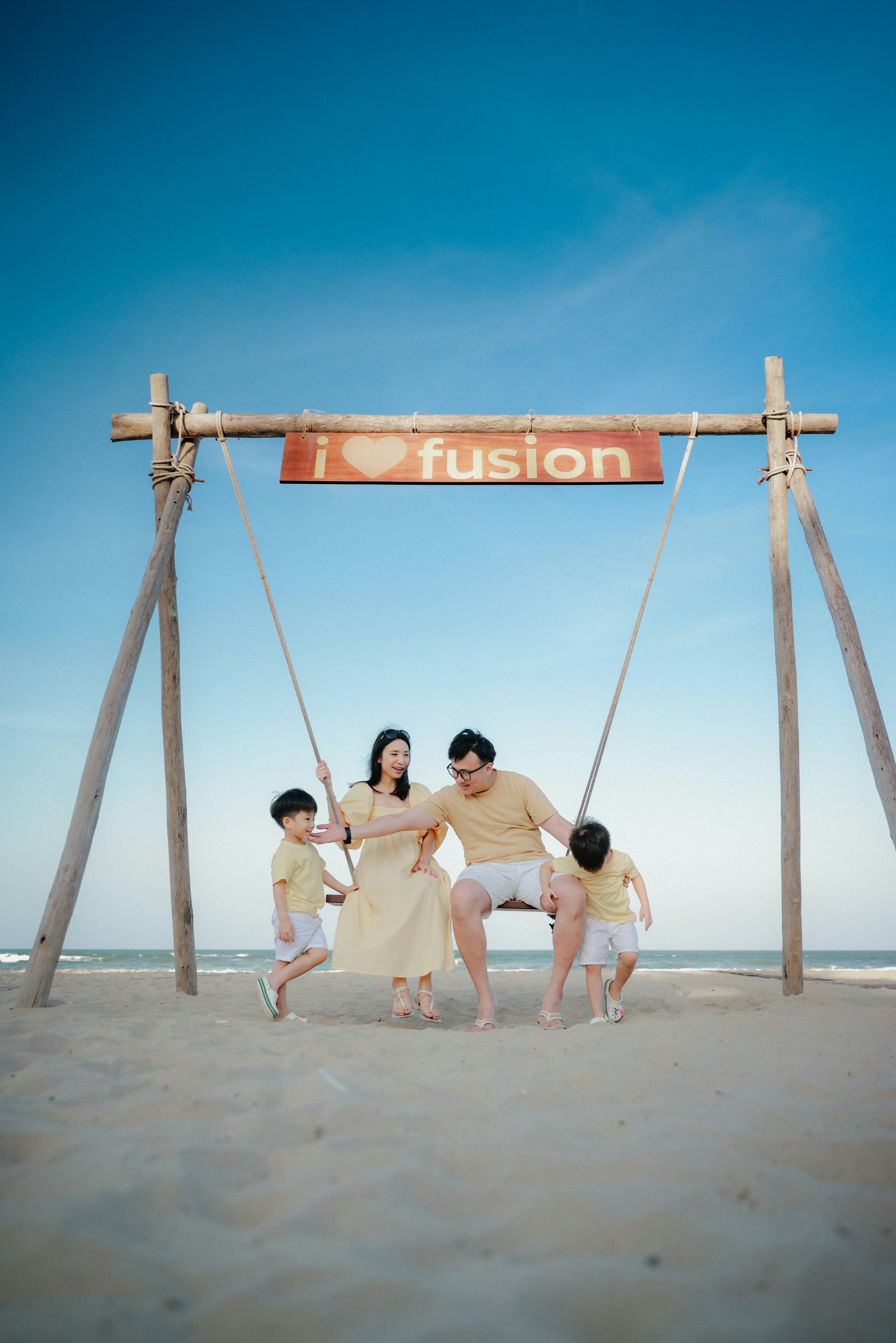 Family on a sunlit beach swing under a sign that reads 'I love fusion'