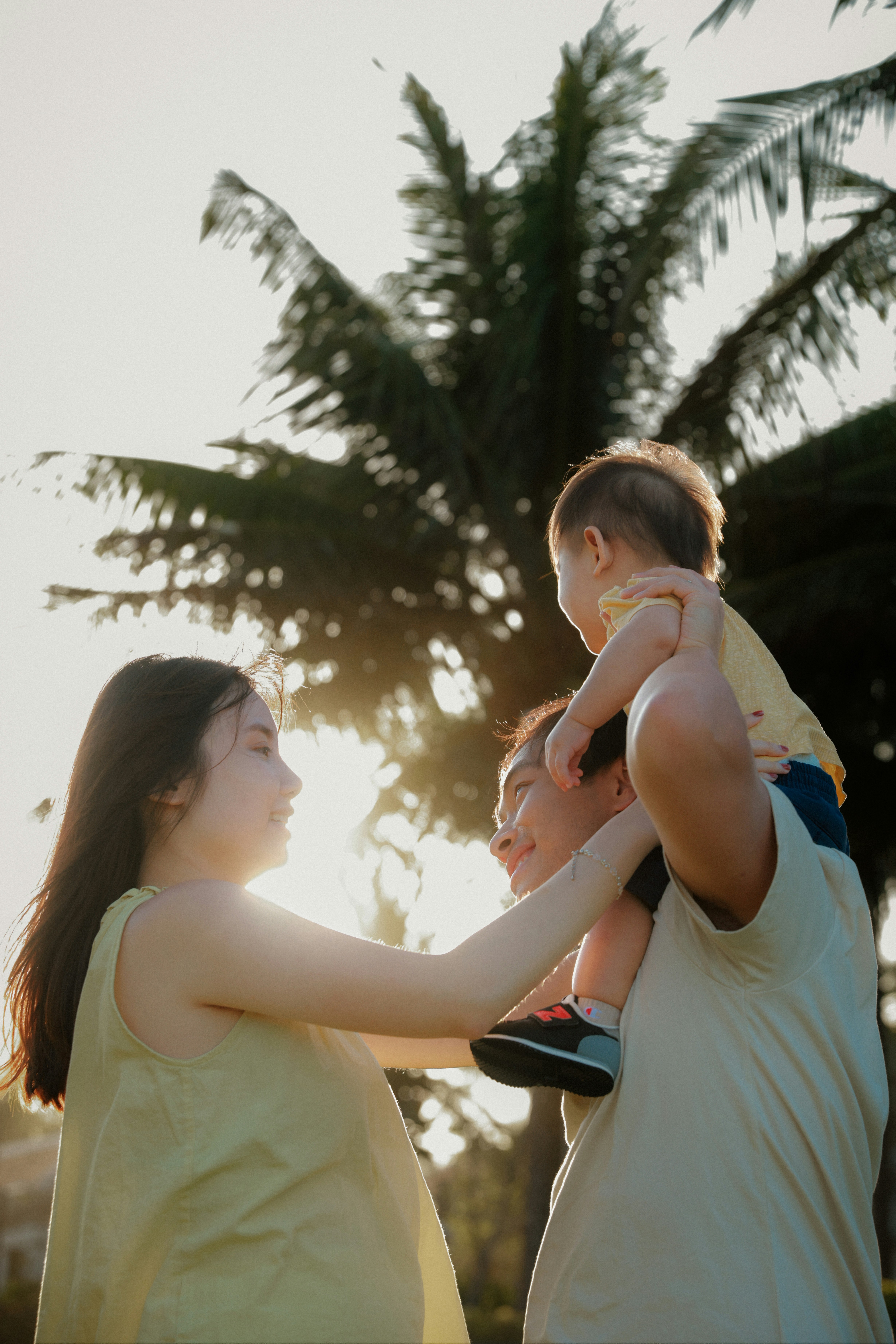 Sunlit Family Moment: A Mother Lifts Her Child as the Sun Glows Behind Palm Trees