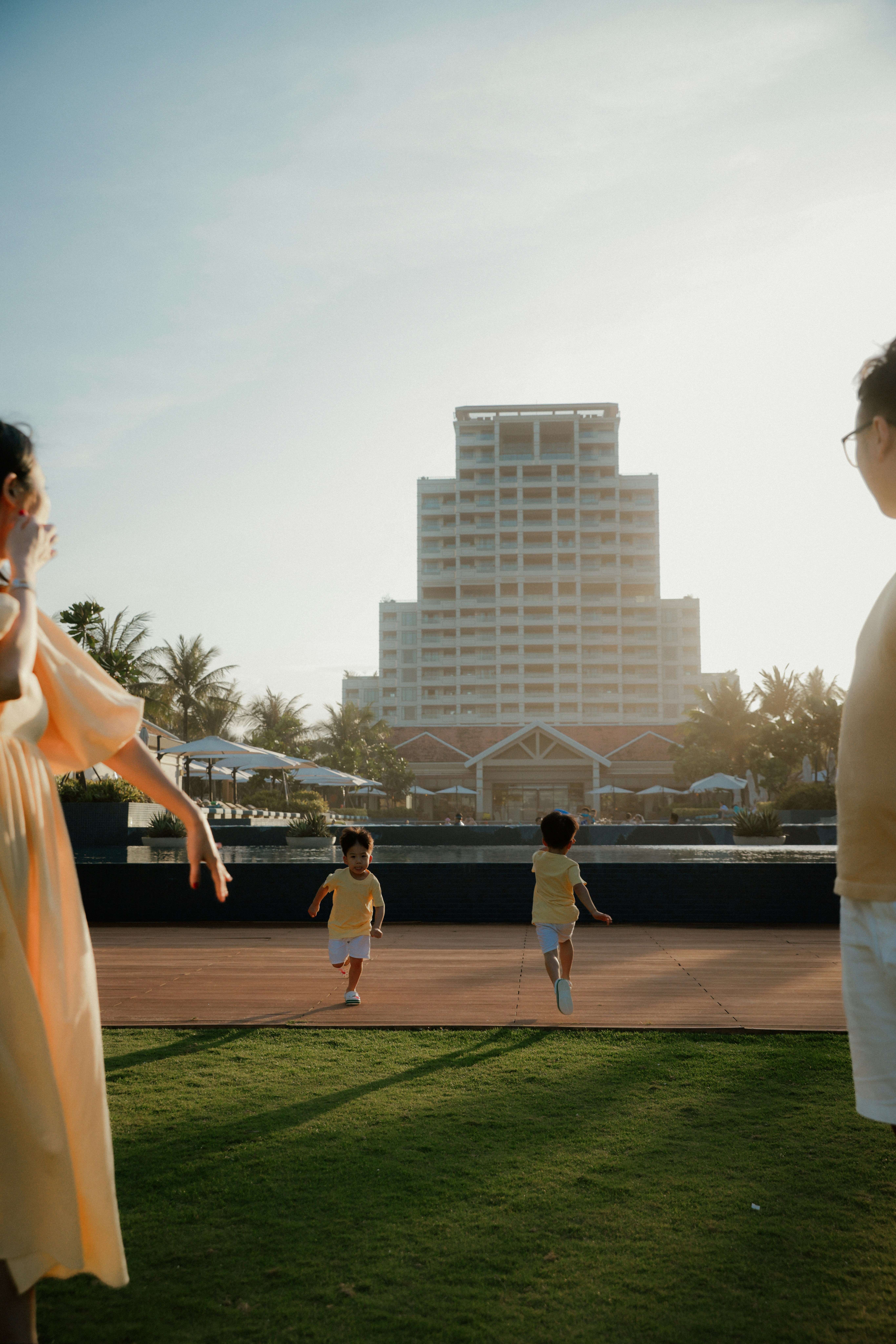 Sunlit sprint: two children dash across a sports field with a modern stepped building in the background