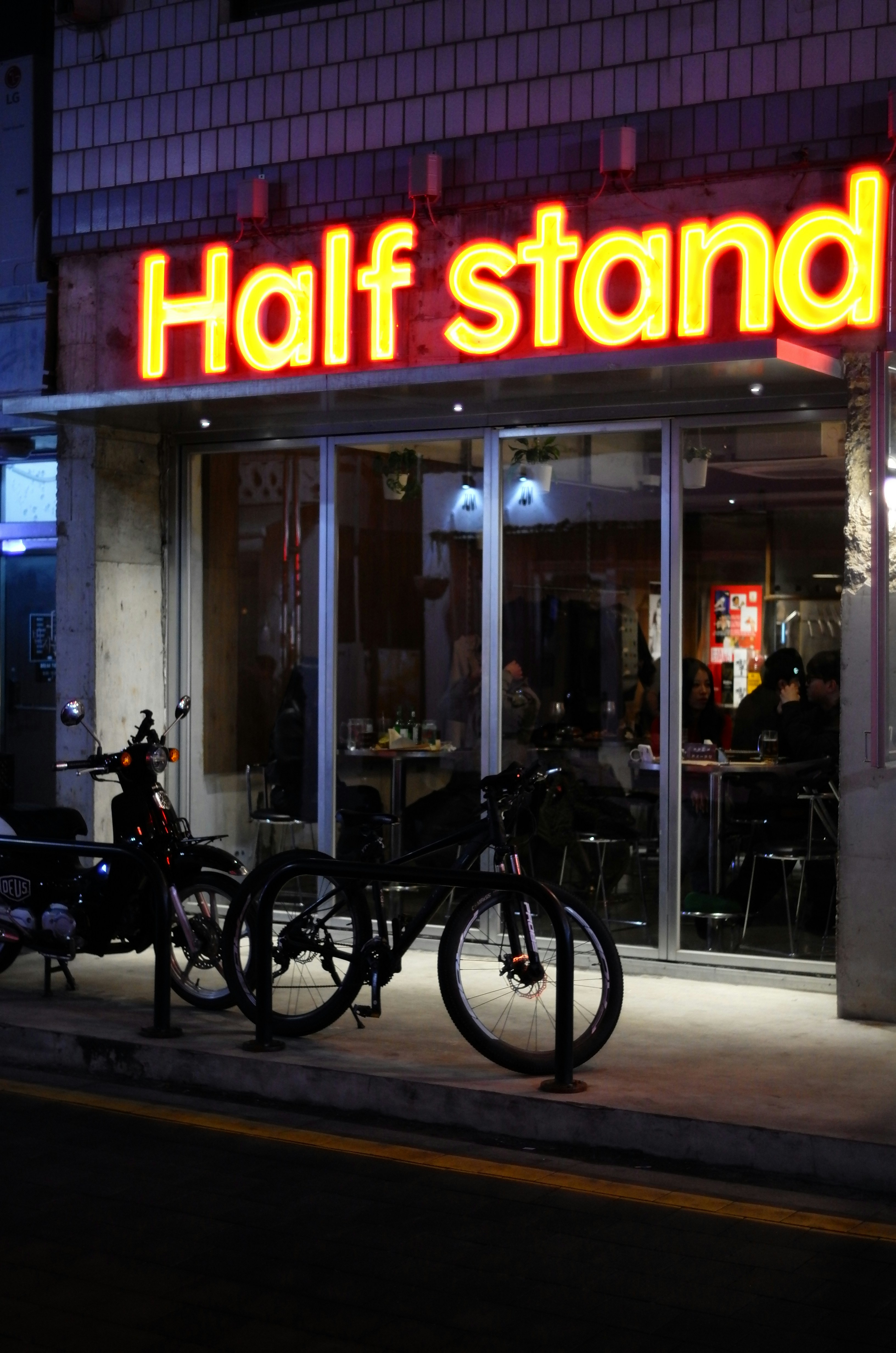 Neon red signage over a glass-front cafe with bicycles parked outside and patrons inside, illuminated storefront at night.