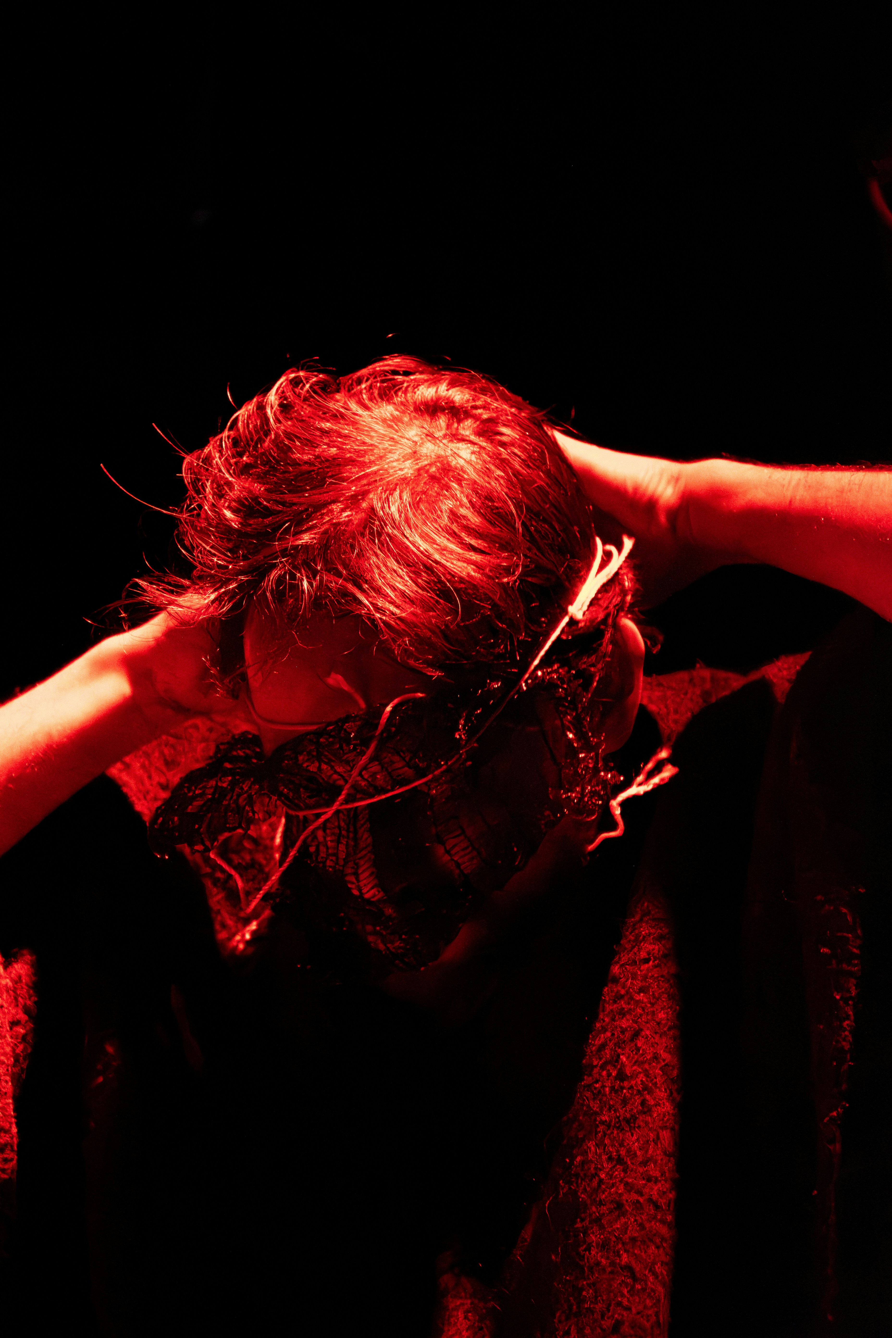 Red-lit silhouette of a performer adjusting a lace mask under dramatic stage lighting