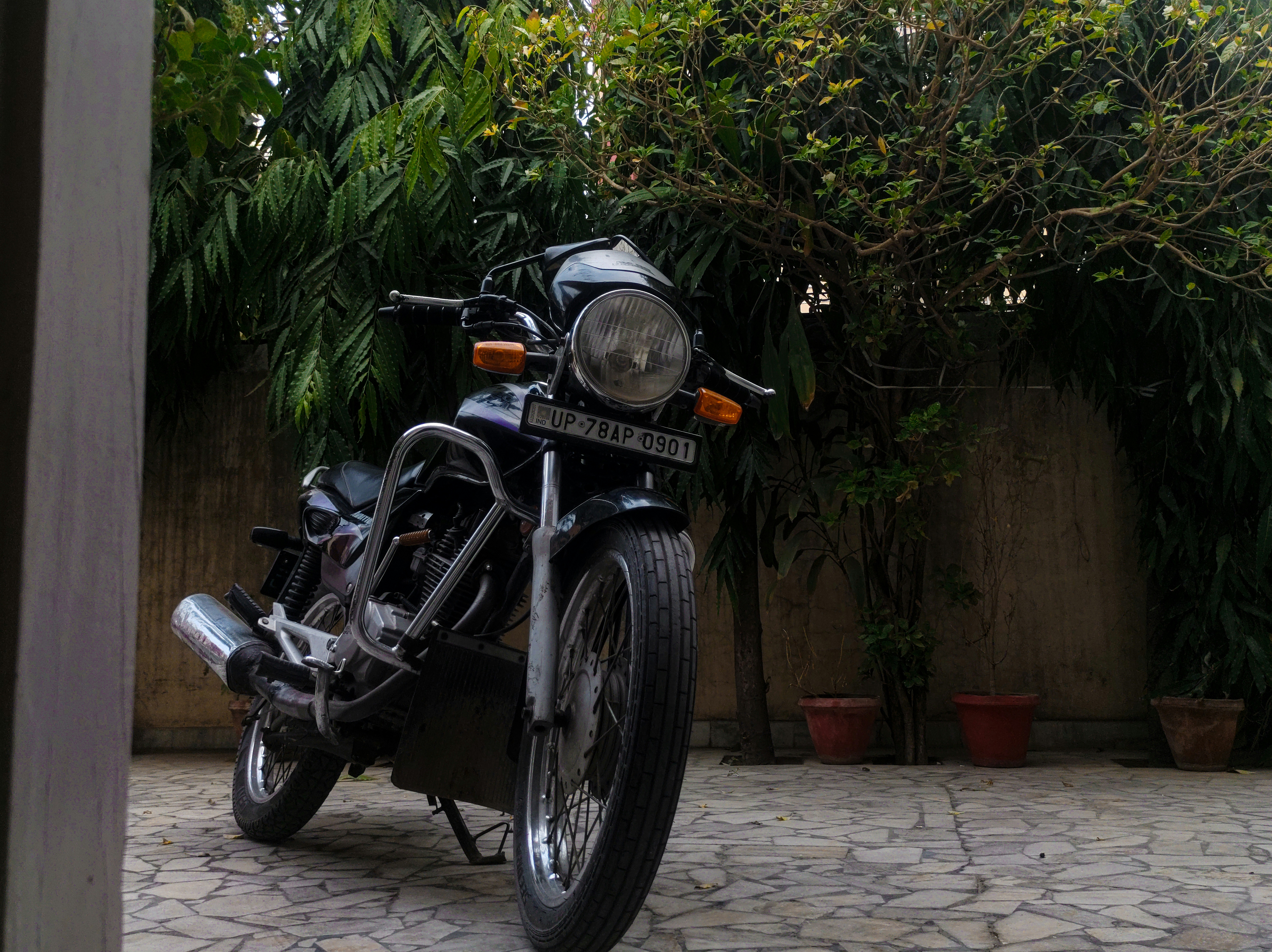 Hero Honda Ambition 133cc 2003 A classic black motorcycle stands parked in a peaceful courtyard, framed by lush green trees and a rustic boundary wall. The low-angle front shot highlights the round headlight, chrome crash guard, and spoke wheels, capturing the raw character of everyday Indian street life. Shot in natural diffused light with a moody, cinematic feel.