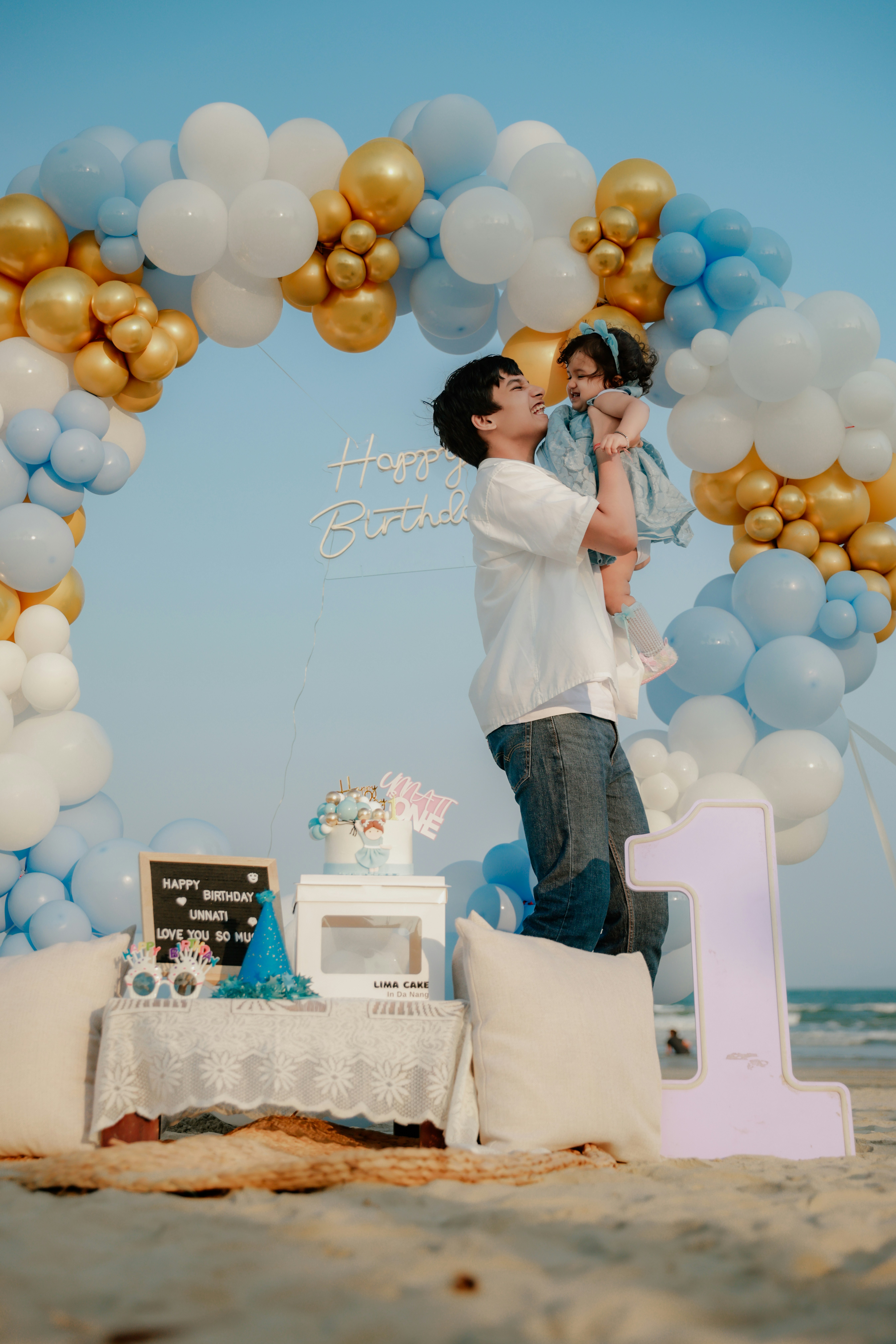 Dad lifts daughter beneath a pastel balloon arch at a seaside birthday setup