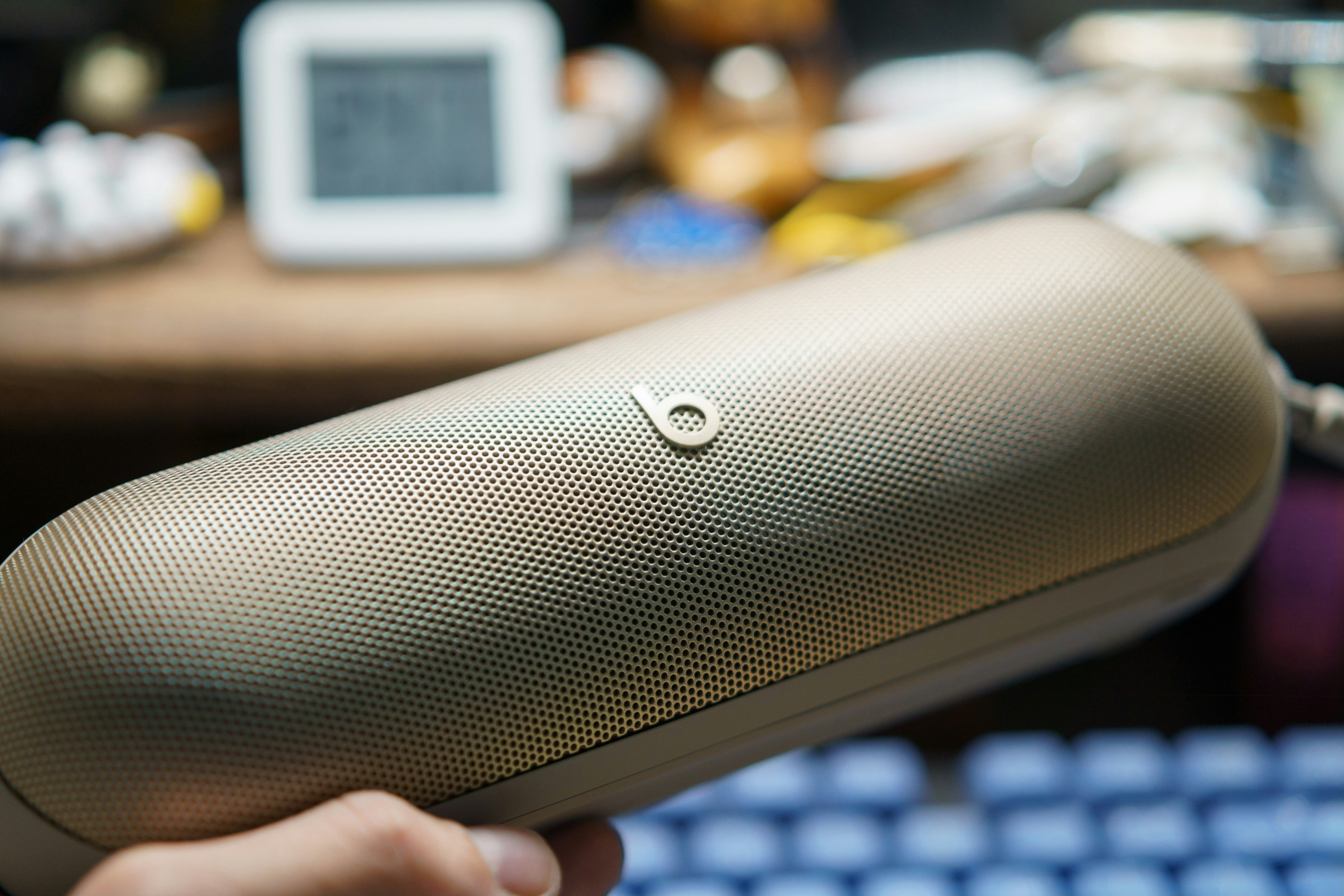 A Beats portable speaker held in the foreground with a blurred desk setup in the background; the focus is on the perforated metal surface and logo, highlighting product texture. logo indicates a consumer electronics device.