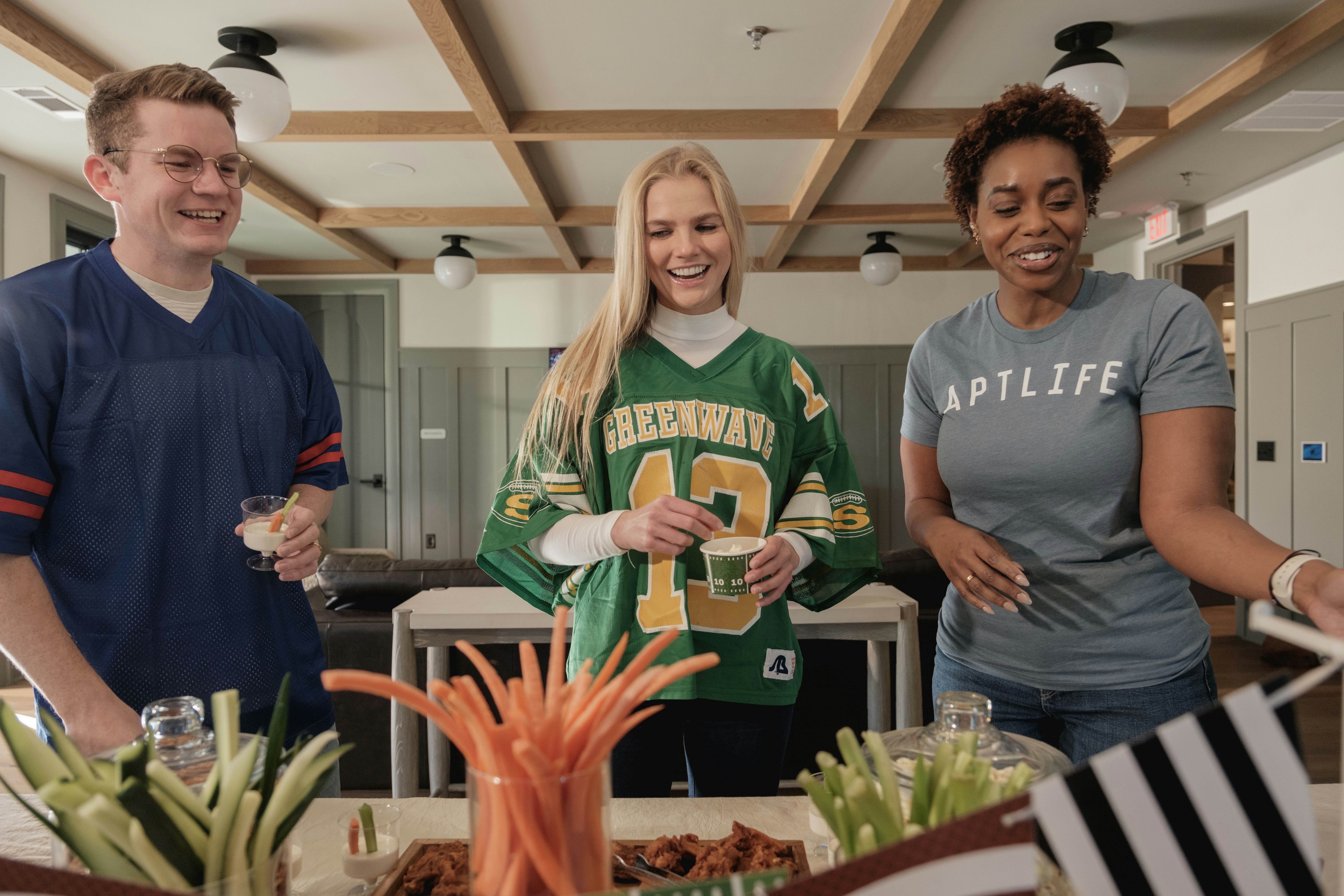 Three friends share a cheerful moment around a buffet at a casual gathering, with greenery and flags in the foreground.