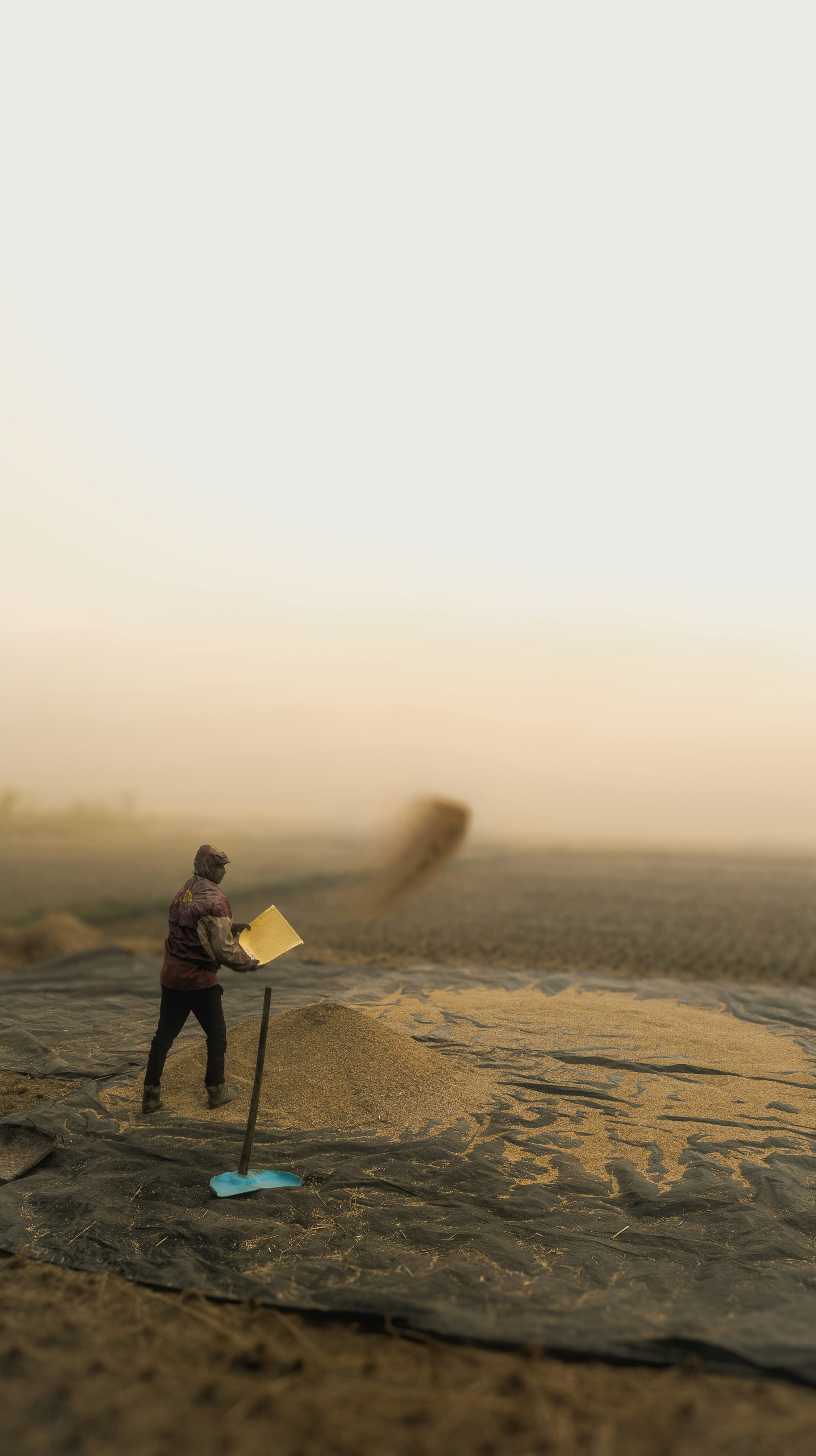 A solitary figure in a hooded jacket surveys a sandy, wind-scoured landscape with a map, standing near a small stake on a tarped ground; a faint haze blanks the horizon, suggesting dusk or sandstorms. This is a staged documentary scene.