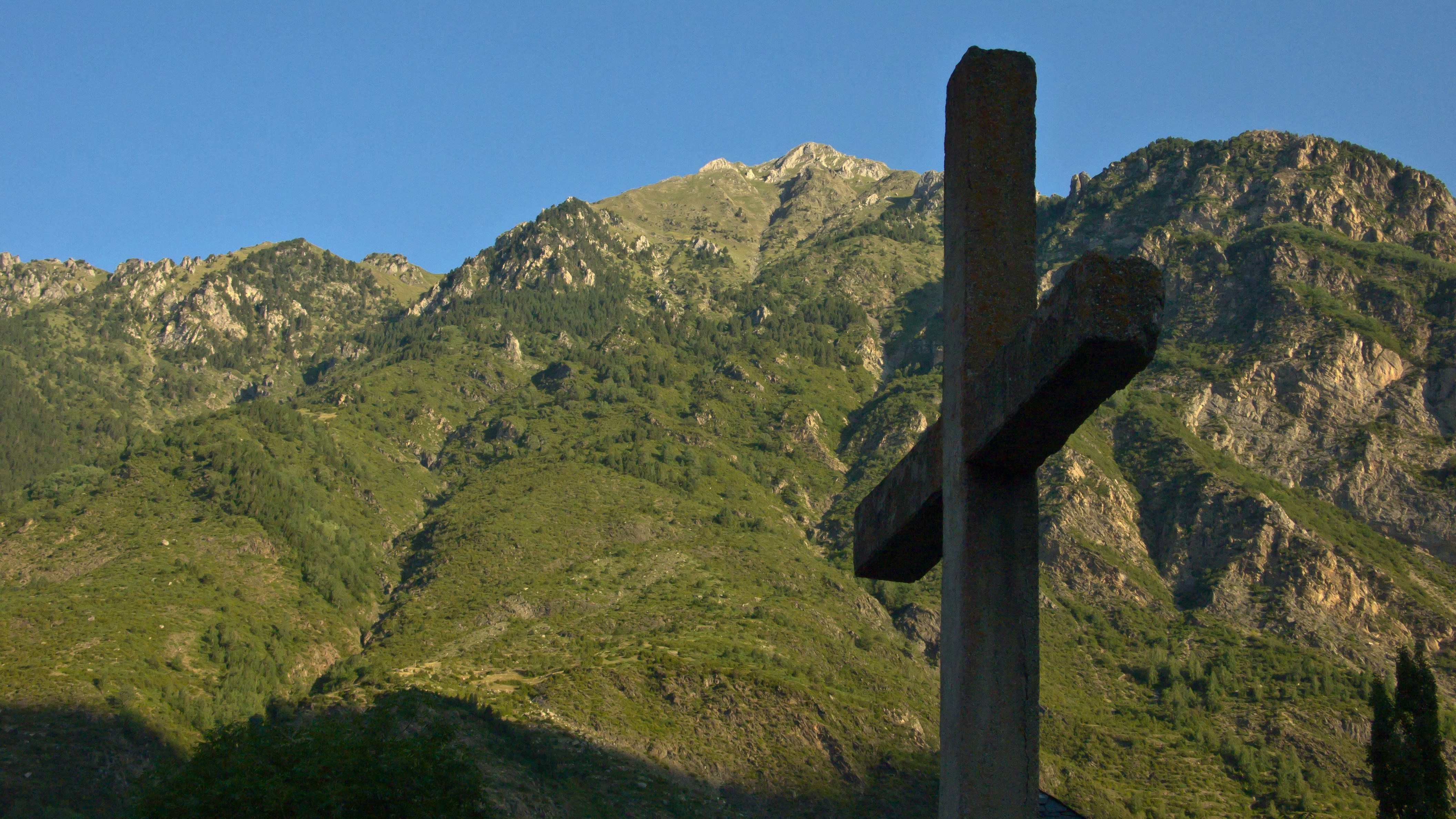 A tall stone cross stands in the foreground against a backdrop of steep, green mountains under a clear blue sky, creating a peaceful and contemplative mountain landscape.
