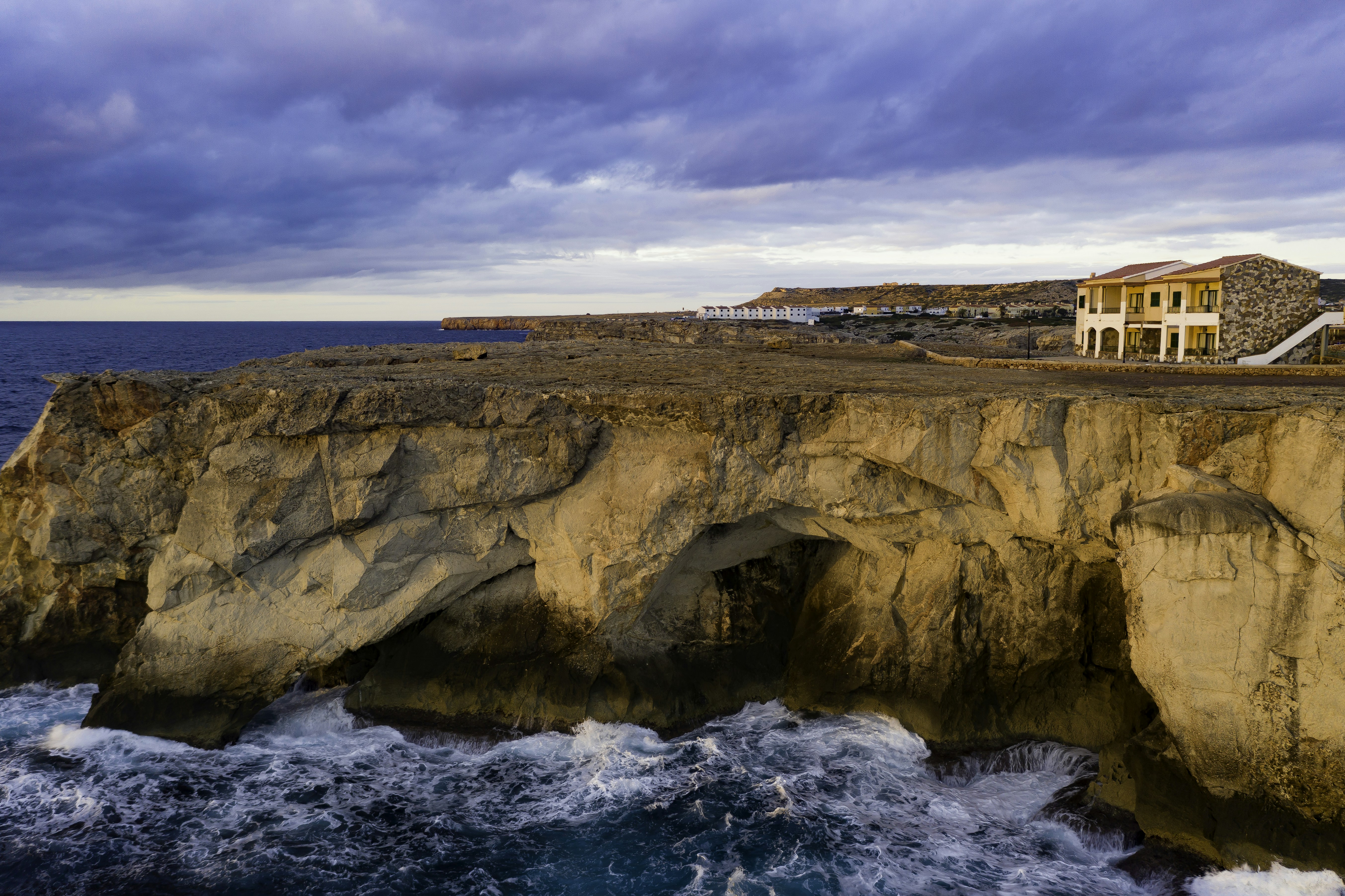 Low coastal cliffs overlook churning blue waters with a distant seaside building, capturing a sunlit edge of land meeting sea. This is a landscape photograph of a rocky shore and cliff-top structures.