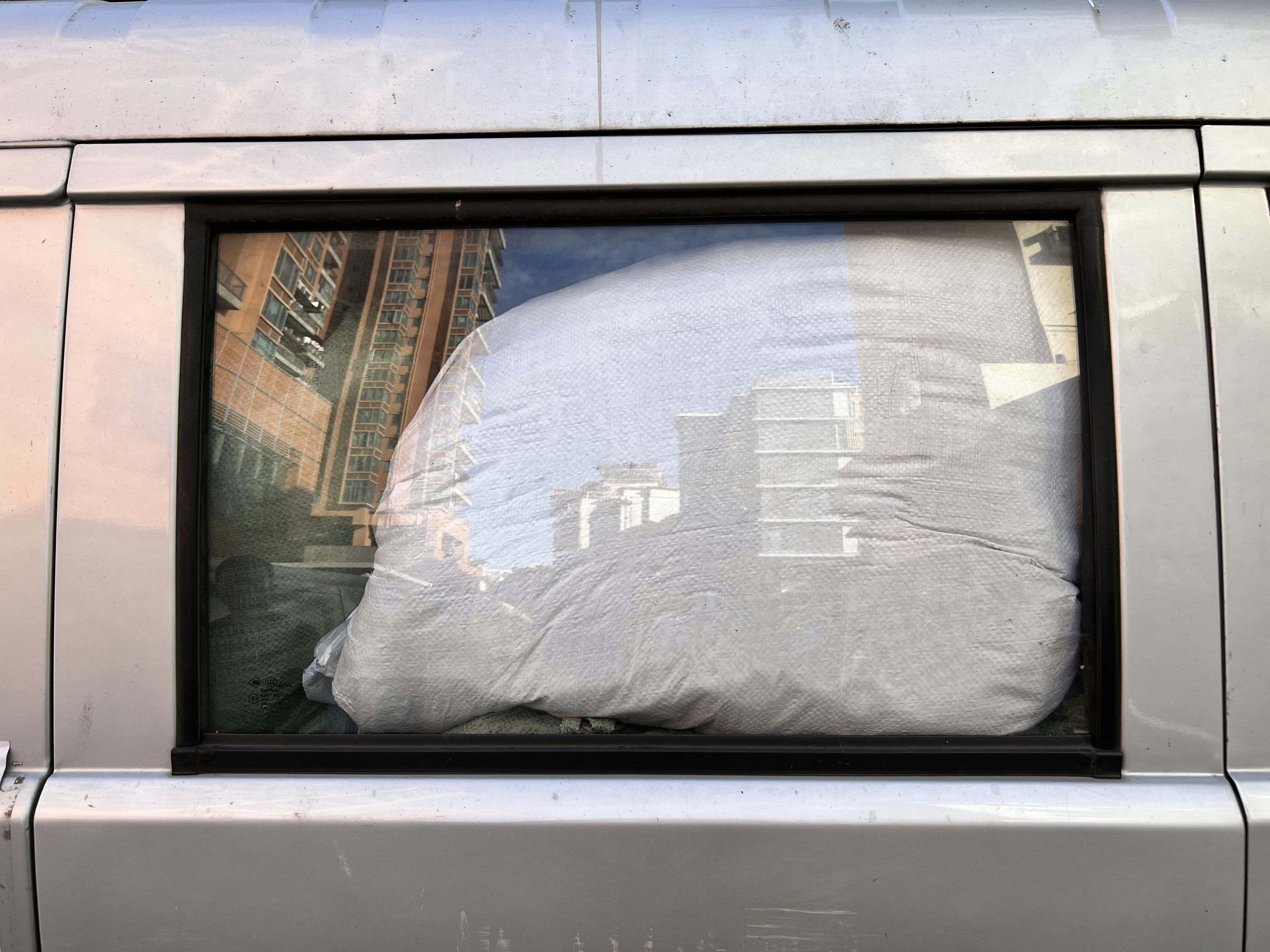 Window reflection showing urban buildings and sky, with a fogged, crumpled fabric cover inside; a car window acts as a mirror-like frame in an urban environment.